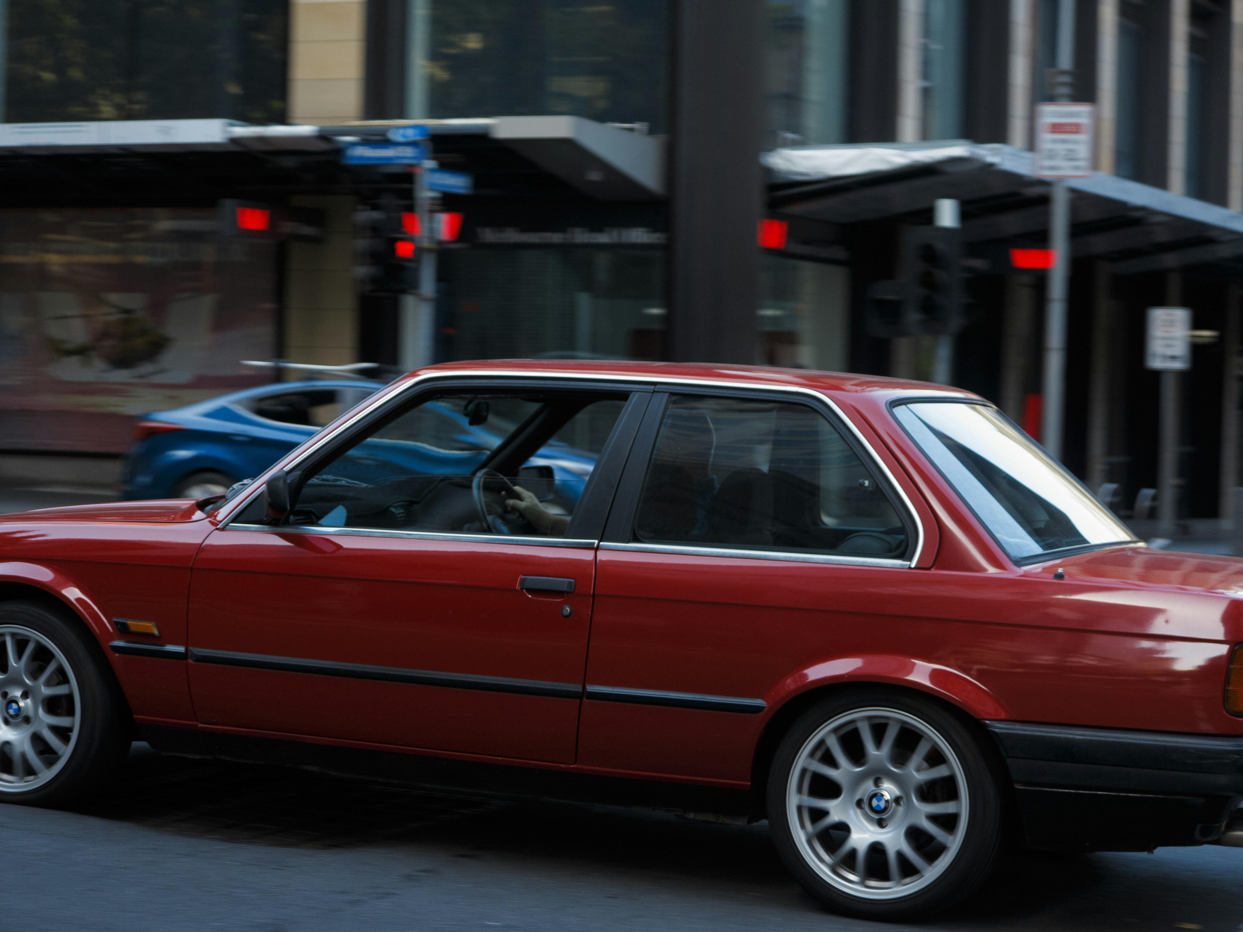 A red classic car navigating through an urban street scene, showcasing its sleek design against modern architecture.
