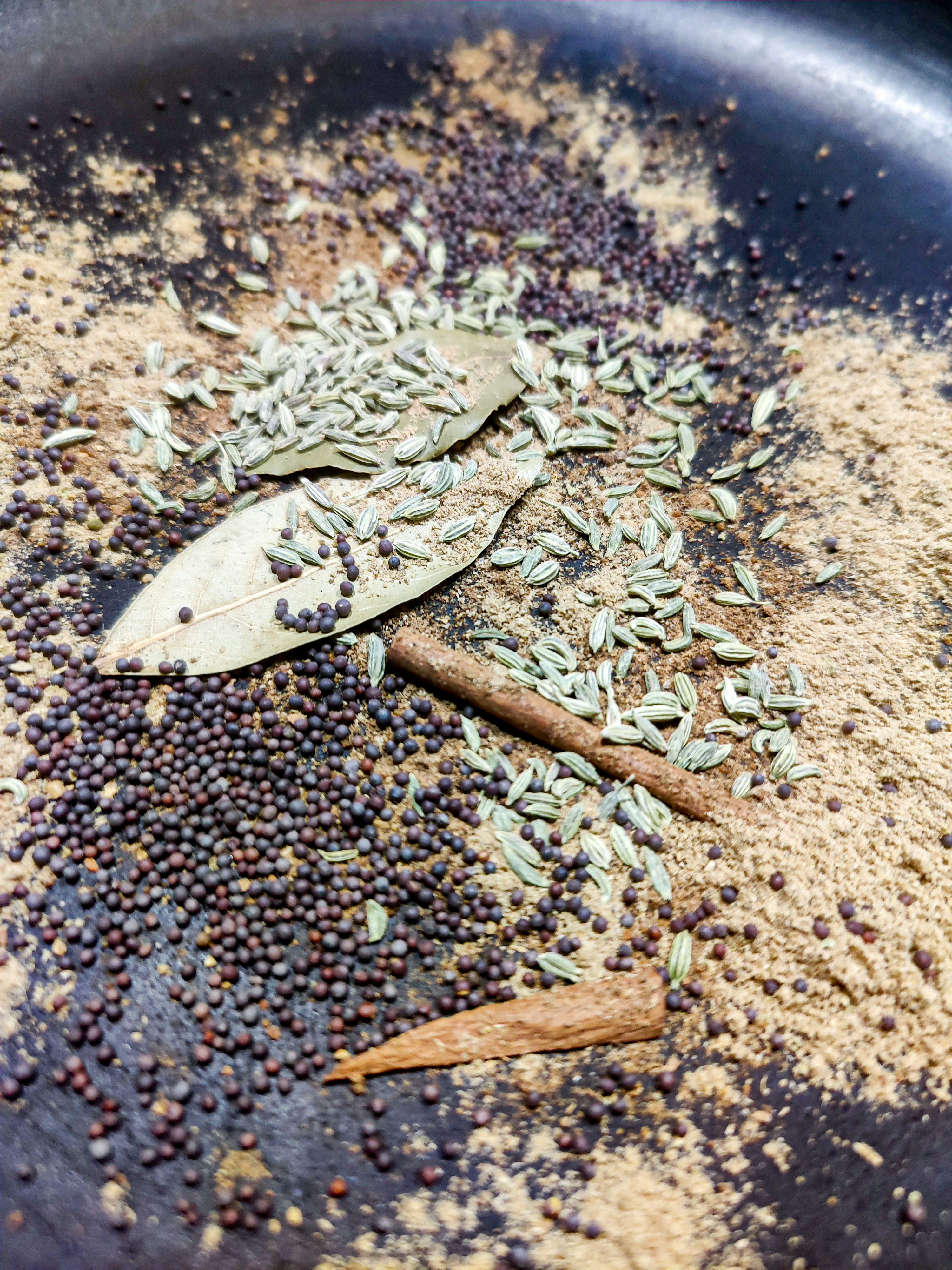 An assortment of spices including mustard seeds, bay leaves, and cinnamon sticks arranged on a dark pan, showcasing their textures and colors.