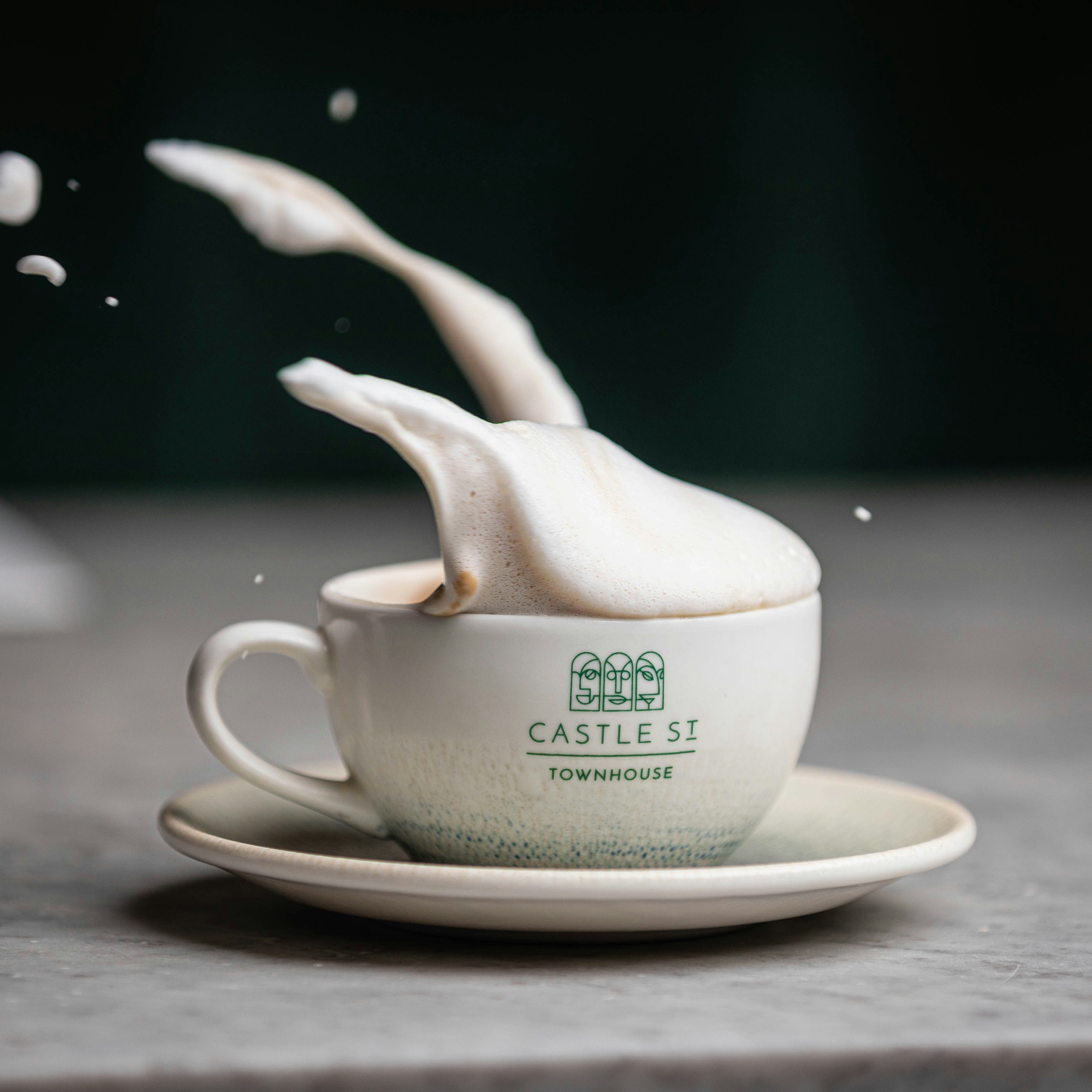 A playful splash of foam erupts from a coffee cup adorned with a logo, set against a softly blurred green background.