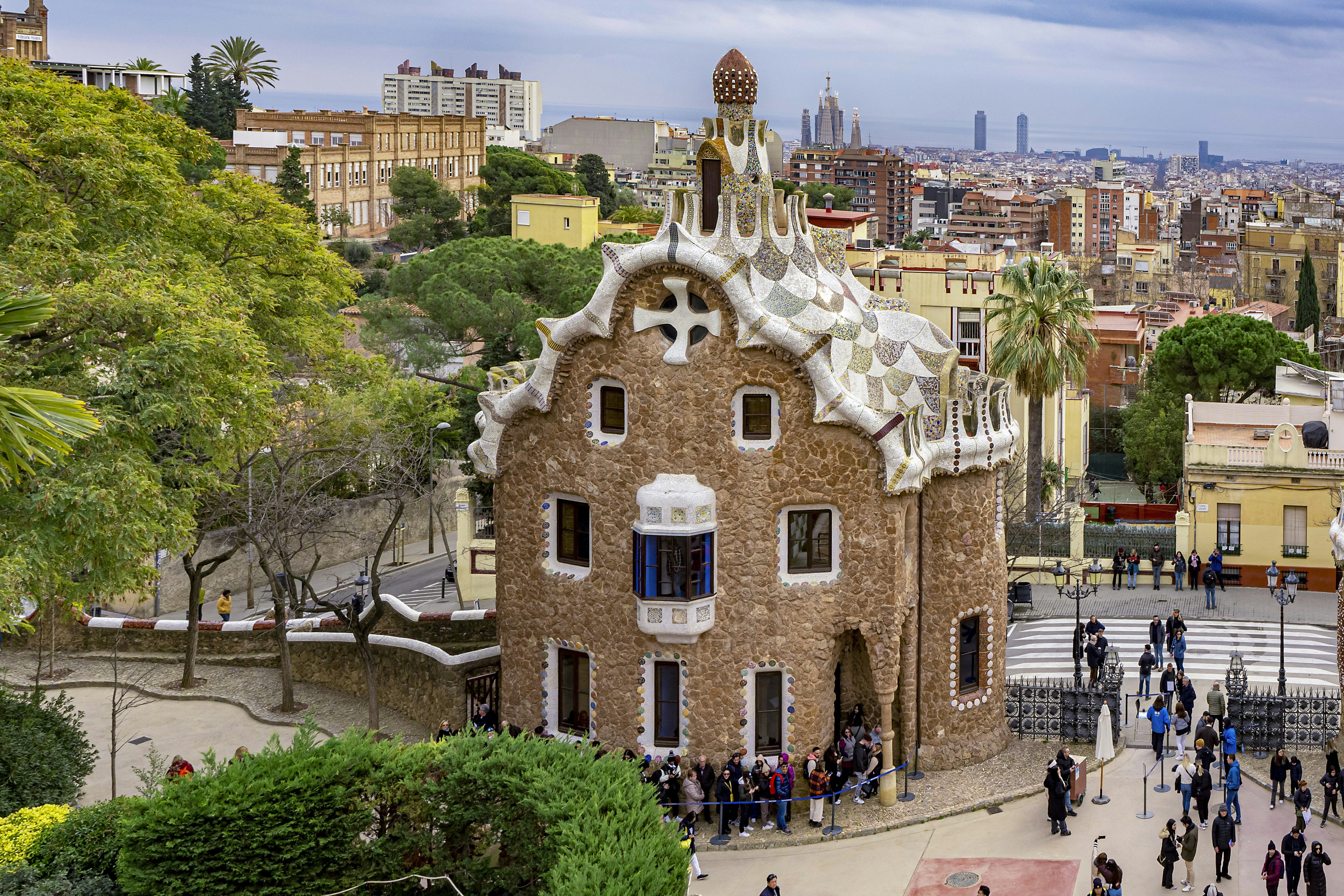 Gaudí-designed building in Park Güell surrounded by lush greenery and cityscape in the background.