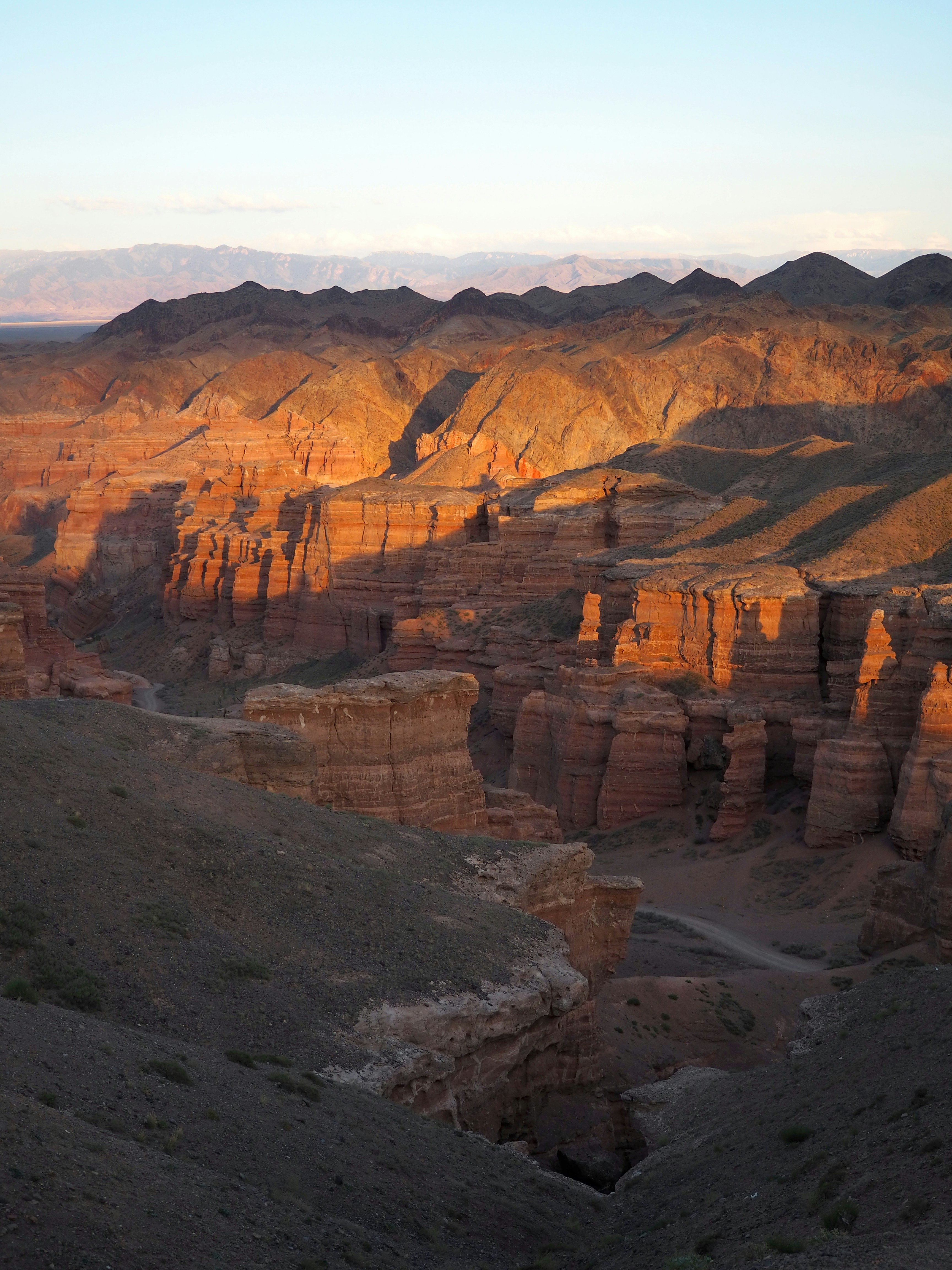 Golden hour sunlight washes layered badlands, lighting orange cliffs and deep crevices across a desert canyon. A wide, tranquil sky overlooks hoodoos and distant mesas in this desert landscape photograph.