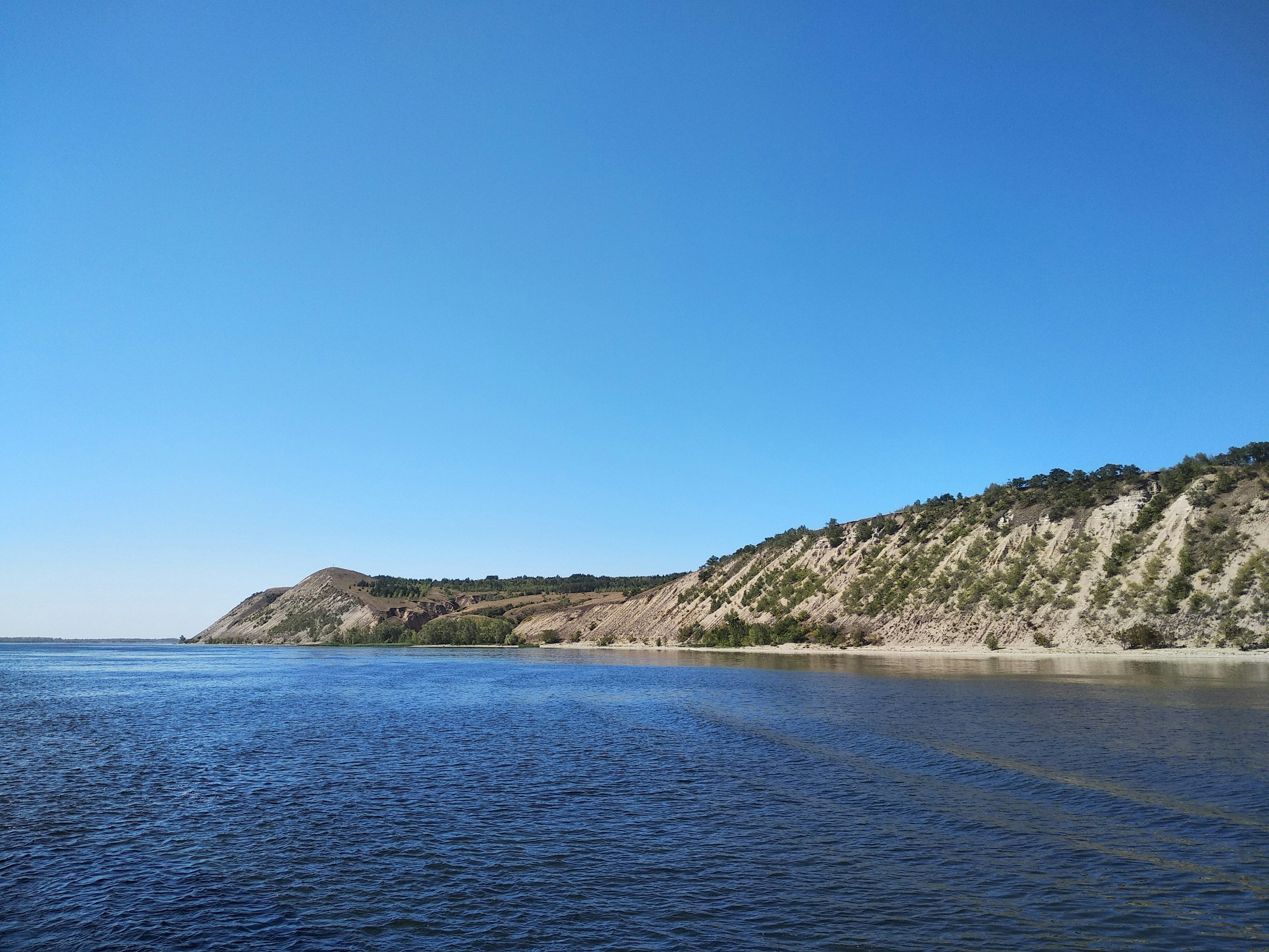 Steep sandy cliffs border a calm river under a clear blue sky.
