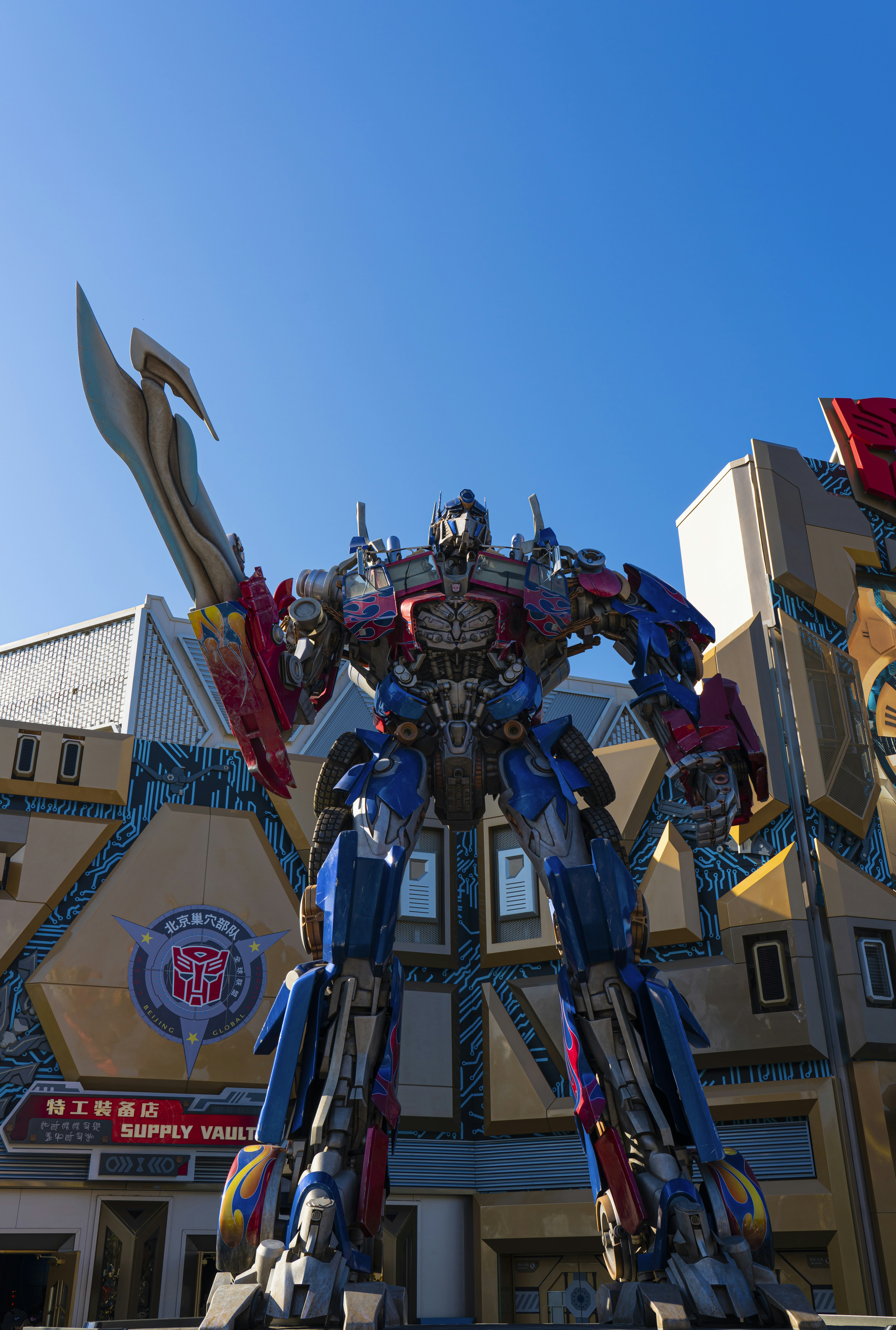 Gigantic robot figure stands proudly against a clear blue sky, showcasing intricate details and vibrant colors. The backdrop features stylized buildings and logos.