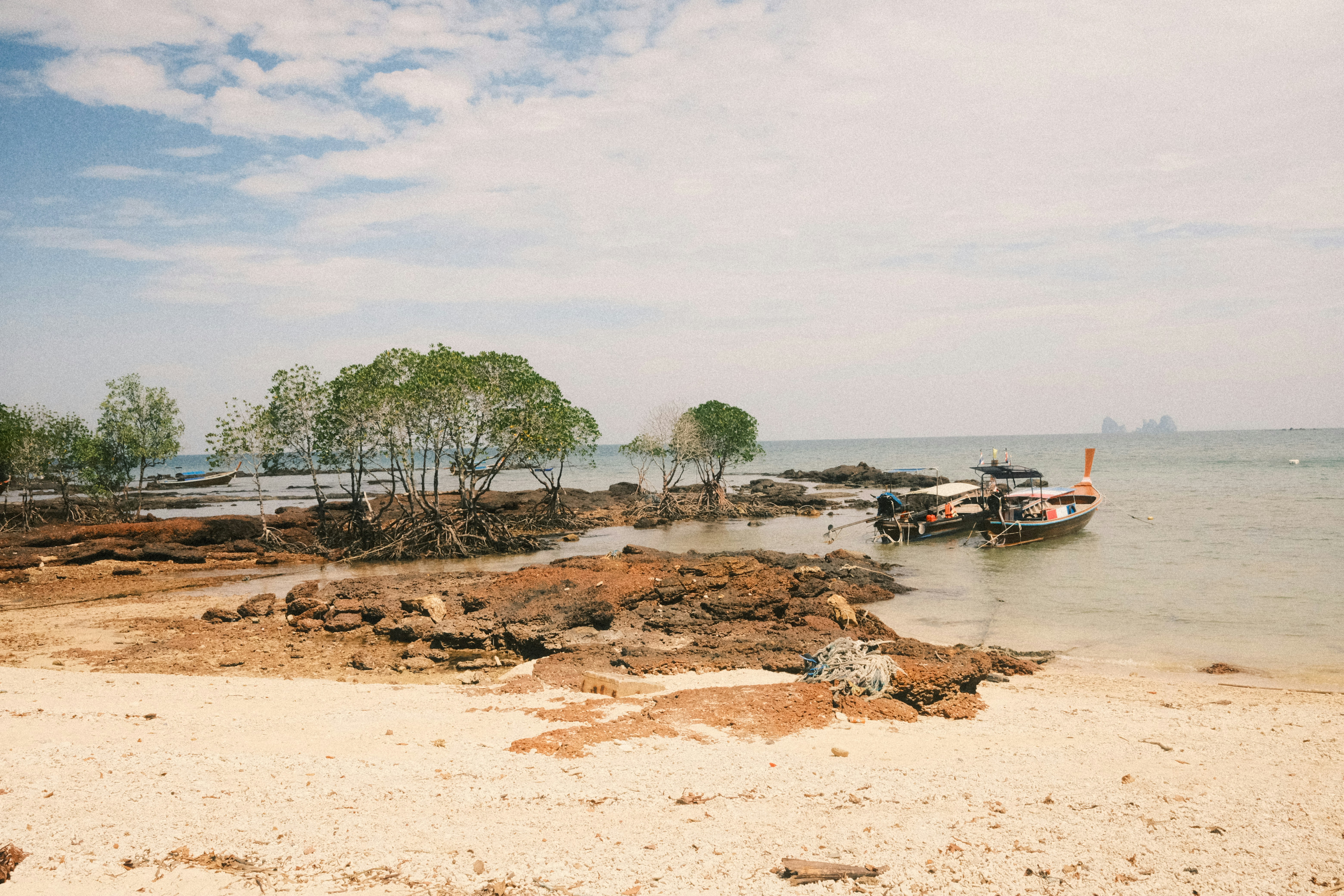 A tranquil beach scene with scattered rocks and a small boat anchored near the shore under a partly cloudy sky.