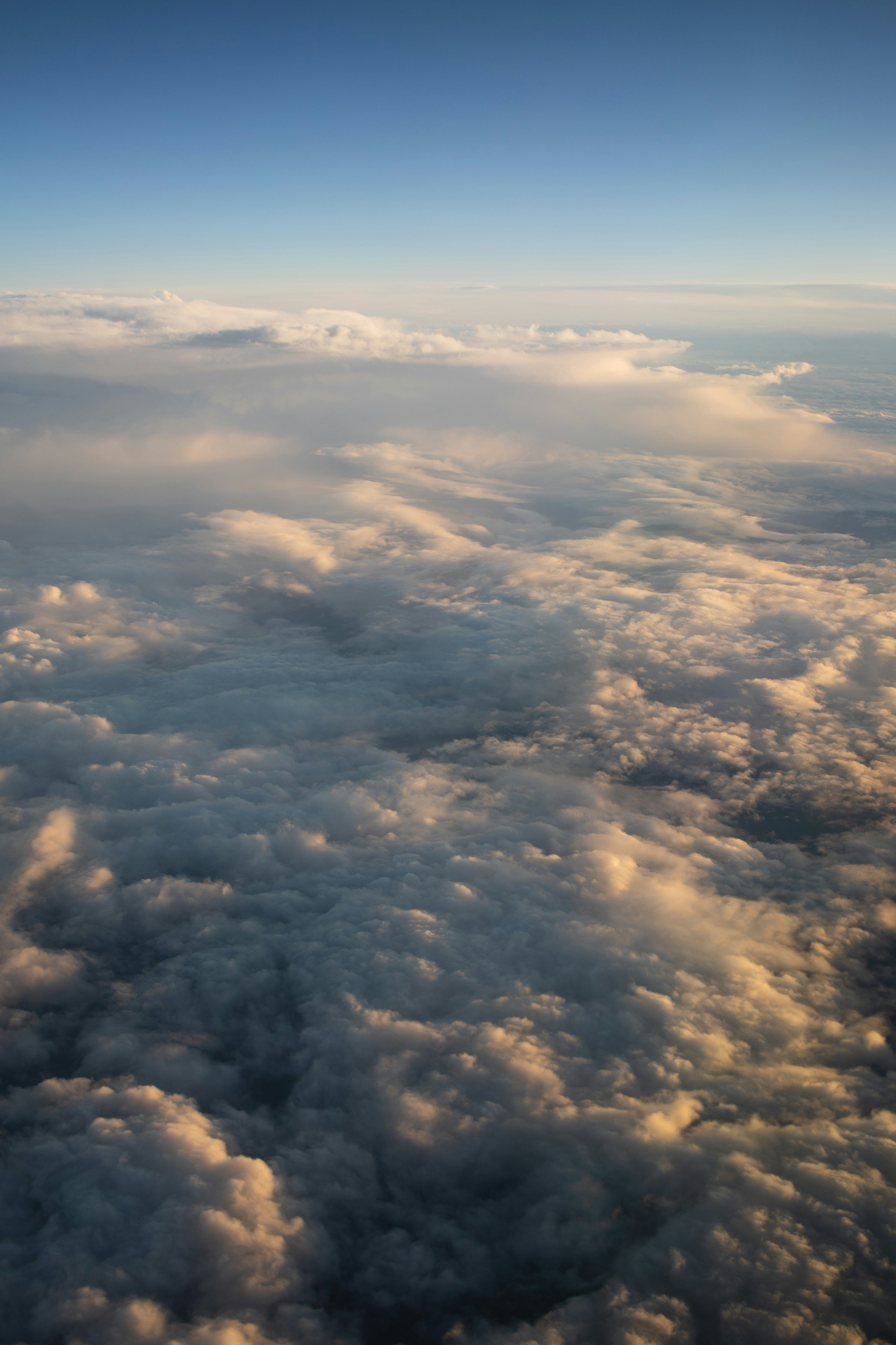 Aerial view of a vast expanse of clouds illuminated by soft, golden light, creating a serene atmosphere. The scene captures the dynamic texture and layers of the cloud formations.
