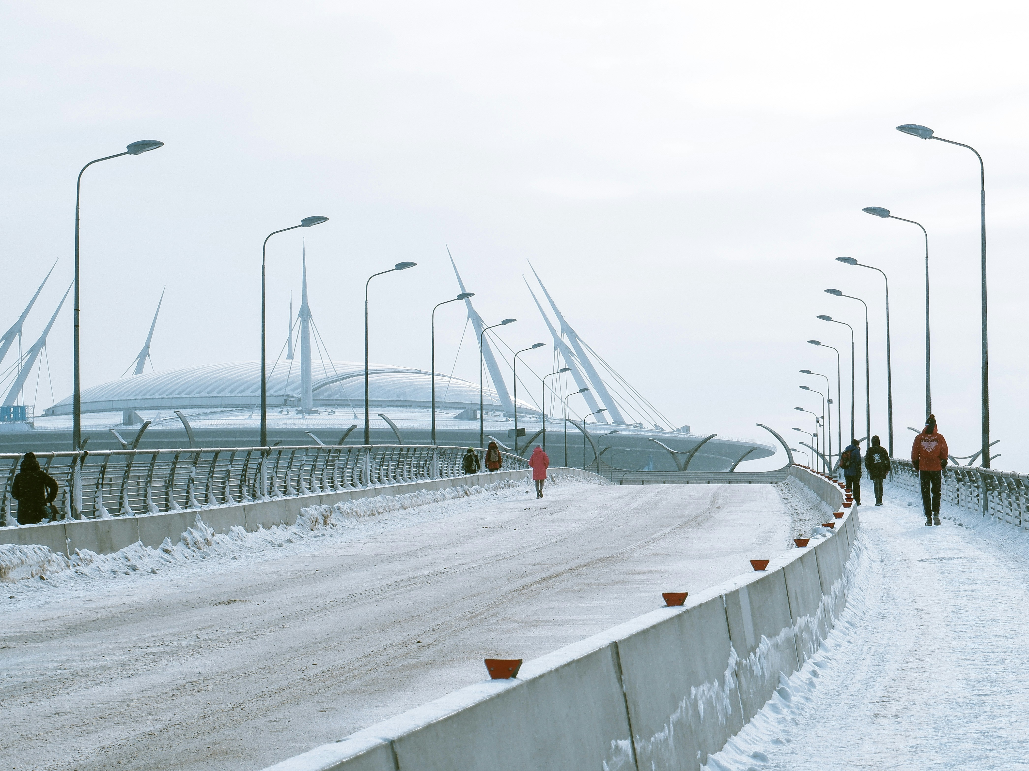 Red-Clad Figure Traverses a Snowbound Bridge Toward a Neo-Futurist Stadium