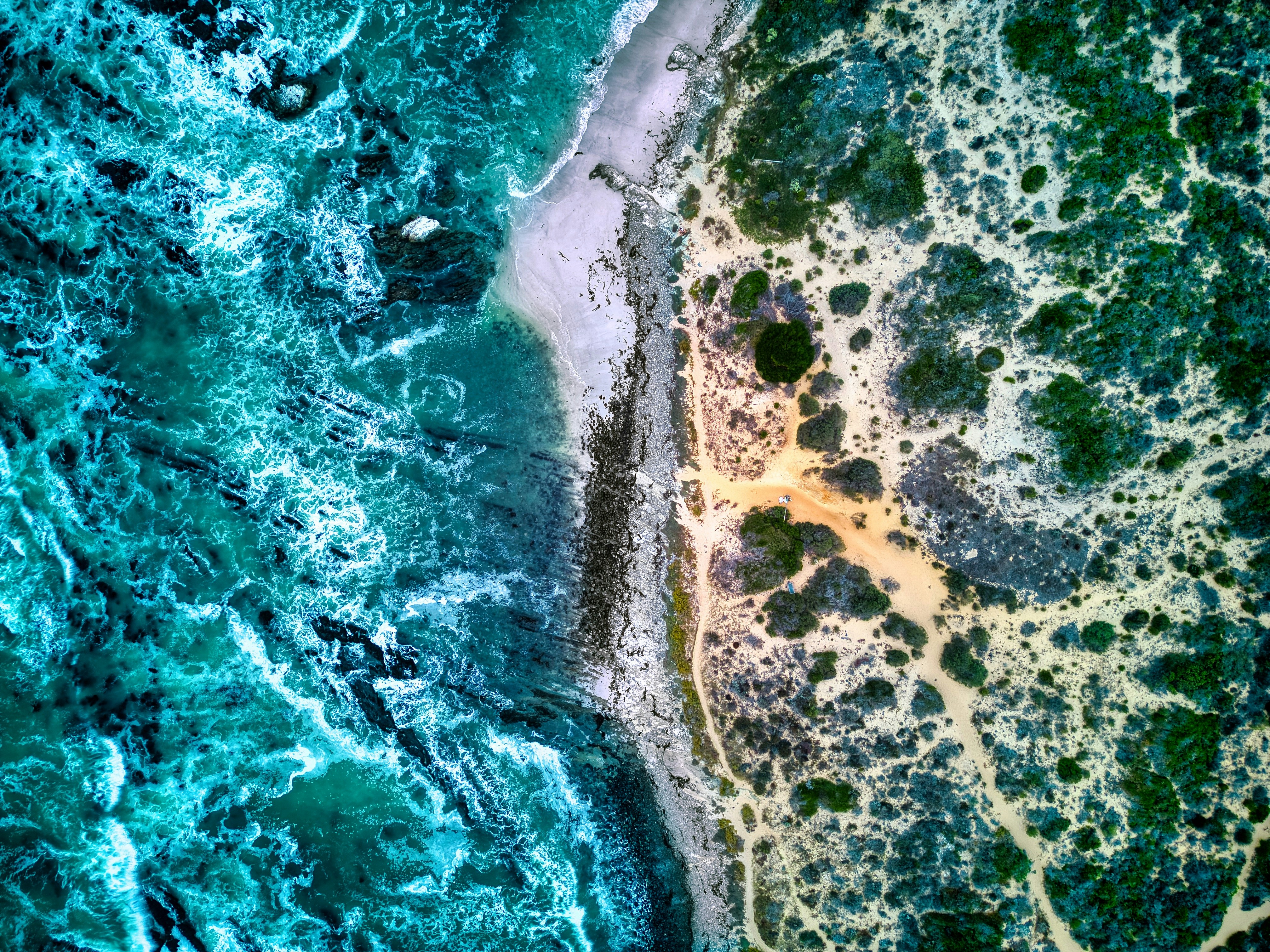 Aerial view of a rugged coastline where vibrant turquoise waves meet a sandy beach, surrounded by rocky terrain and patches of greenery.