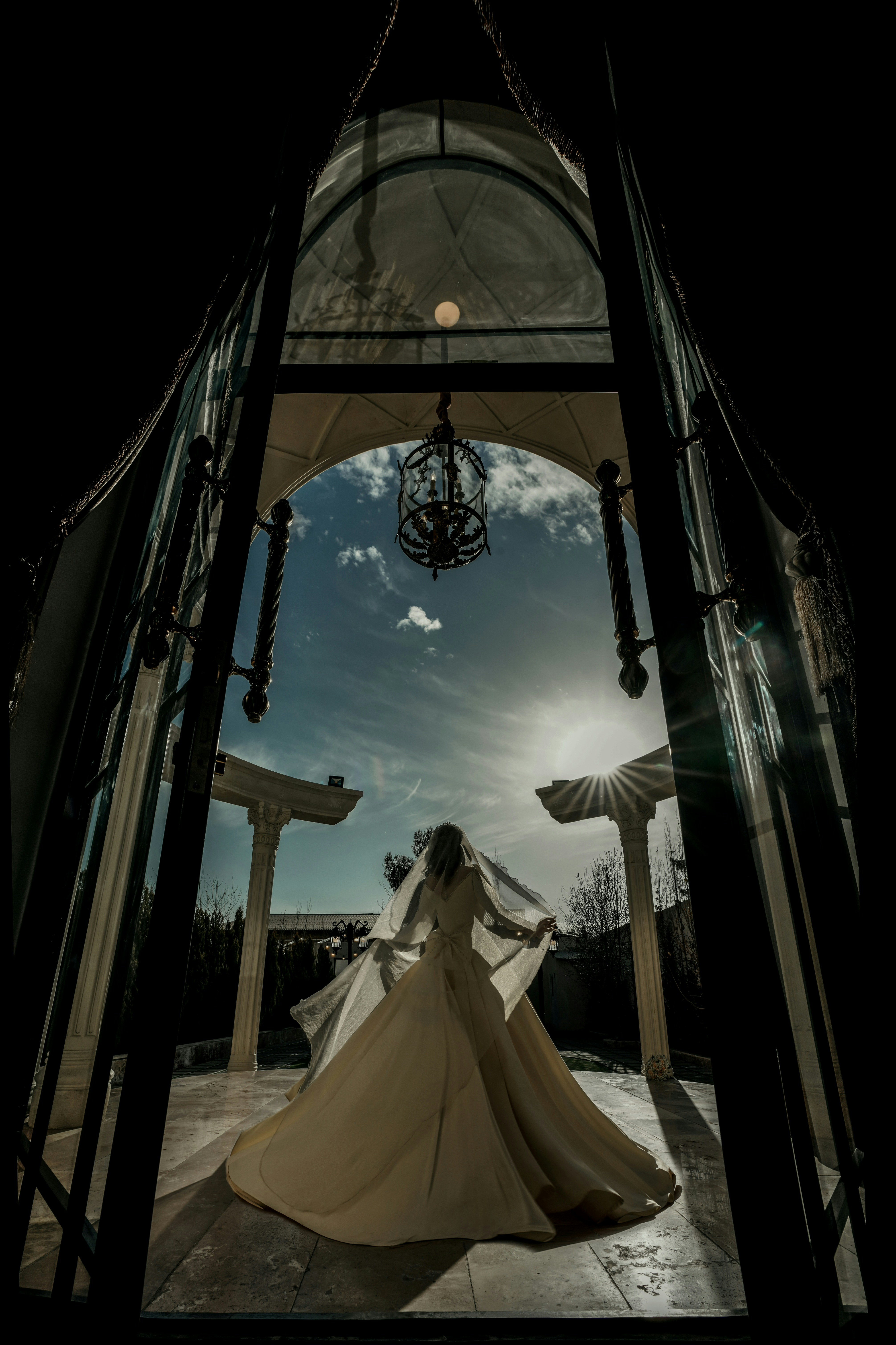 Bride in flowing gown stands gracefully beneath an ornate archway, with sunlight illuminating her veil and the surrounding architecture.
