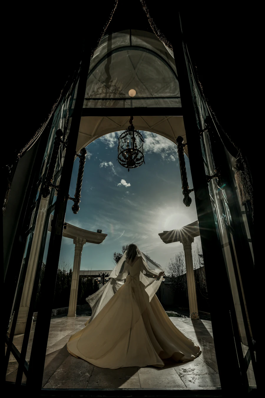 A bride in a flowing gown stands beneath a pavilion, framed by dark doorway edges and columns, with the sun flaring low in the sky behind her. A hanging chandelier and dramatic backlight create a cinematic silhouette.