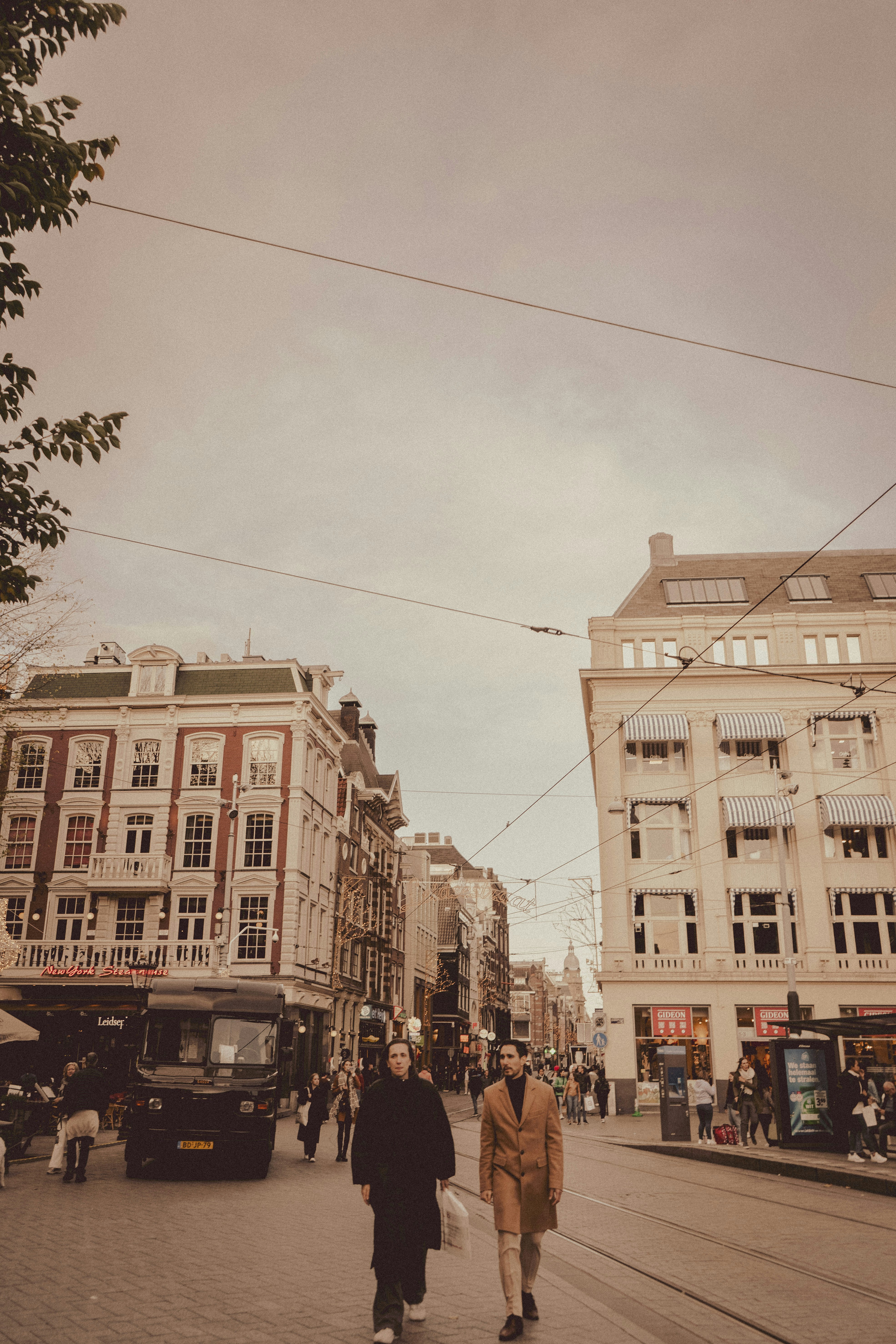 Tan-Coat Duo Under Tram Wires in Sepia Cityscape