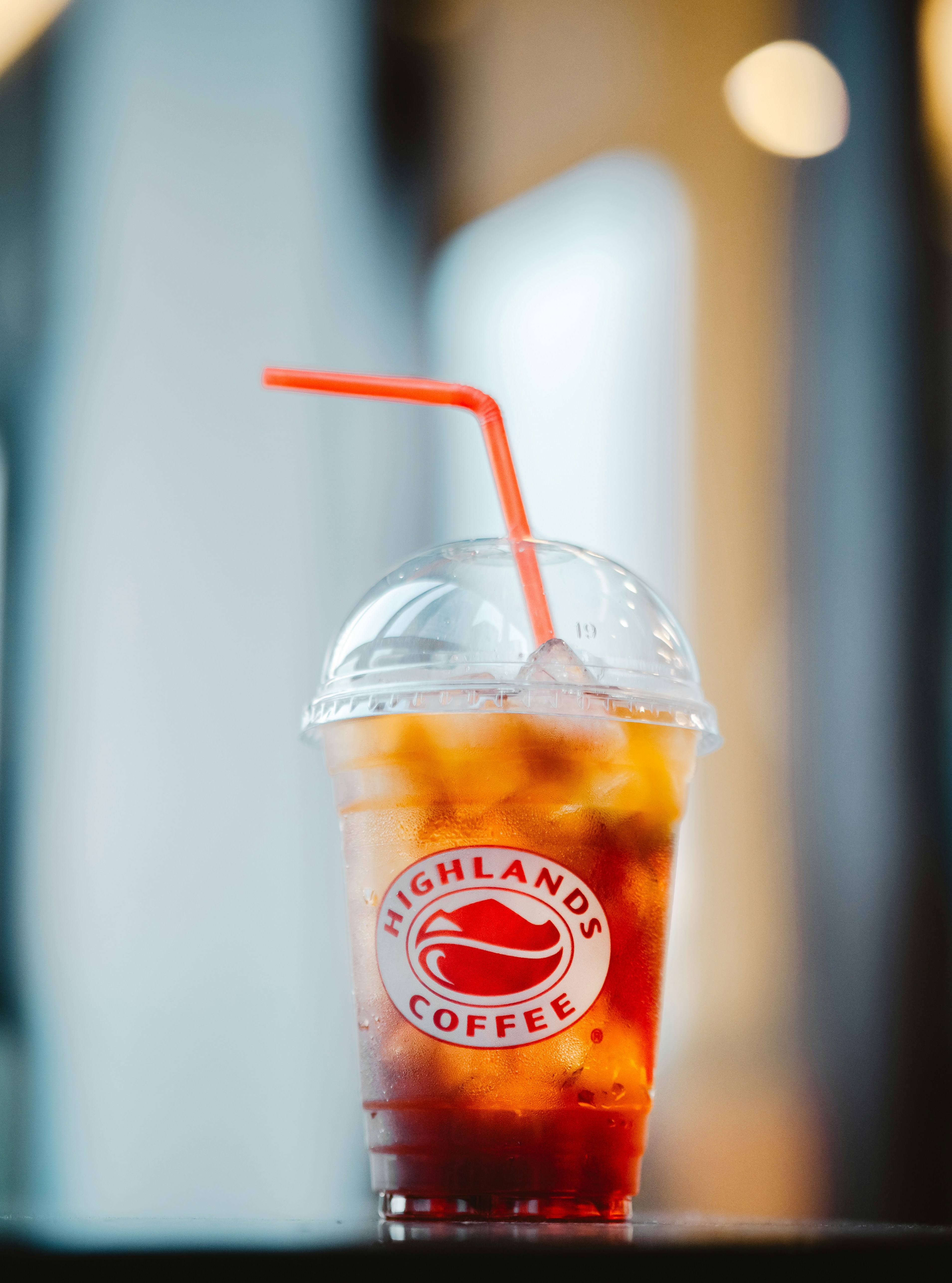 Iced coffee in a clear cup with a dome lid and orange straw bearing the Highlands Coffee logo. Shallow depth of field keeps the drink in crisp focus while the background dissolves into soft cafe bokeh.