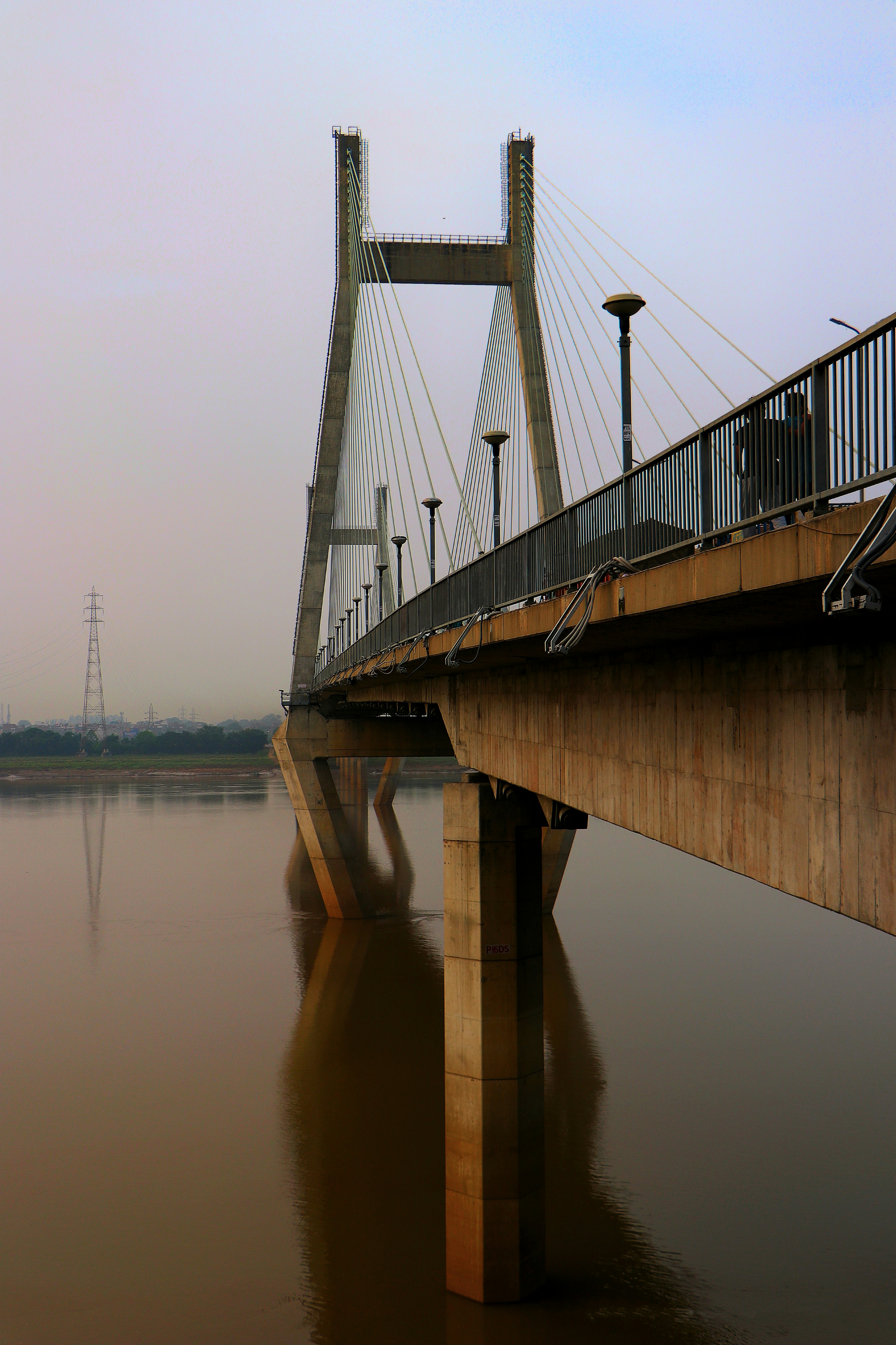 Suspension bridge spanning a calm river, showcasing modern engineering and infrastructure against a muted sky.
