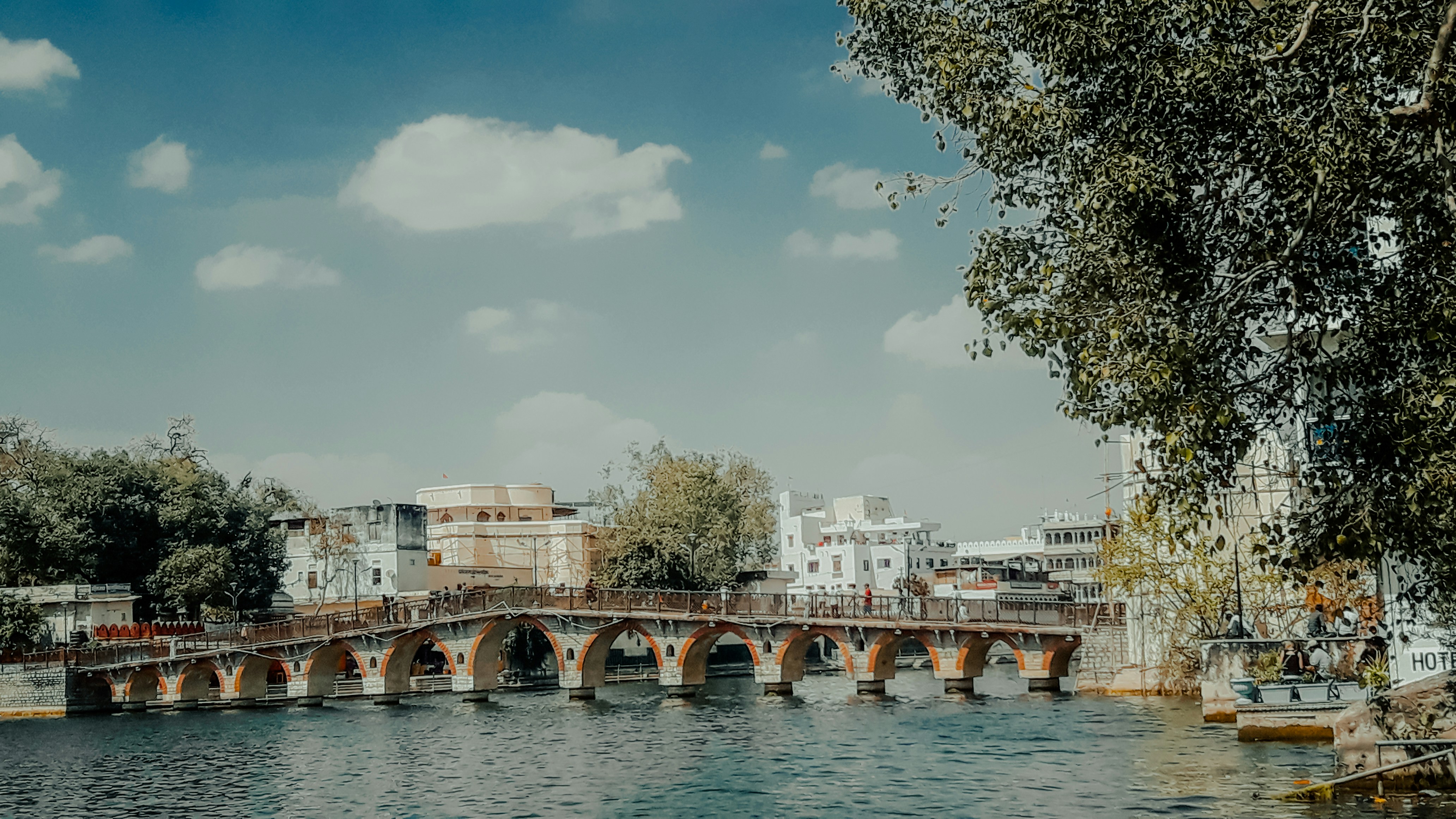 Historic bridge spanning a tranquil river, framed by lush greenery and urban architecture. Clouds drift lazily above, enhancing the serene atmosphere.