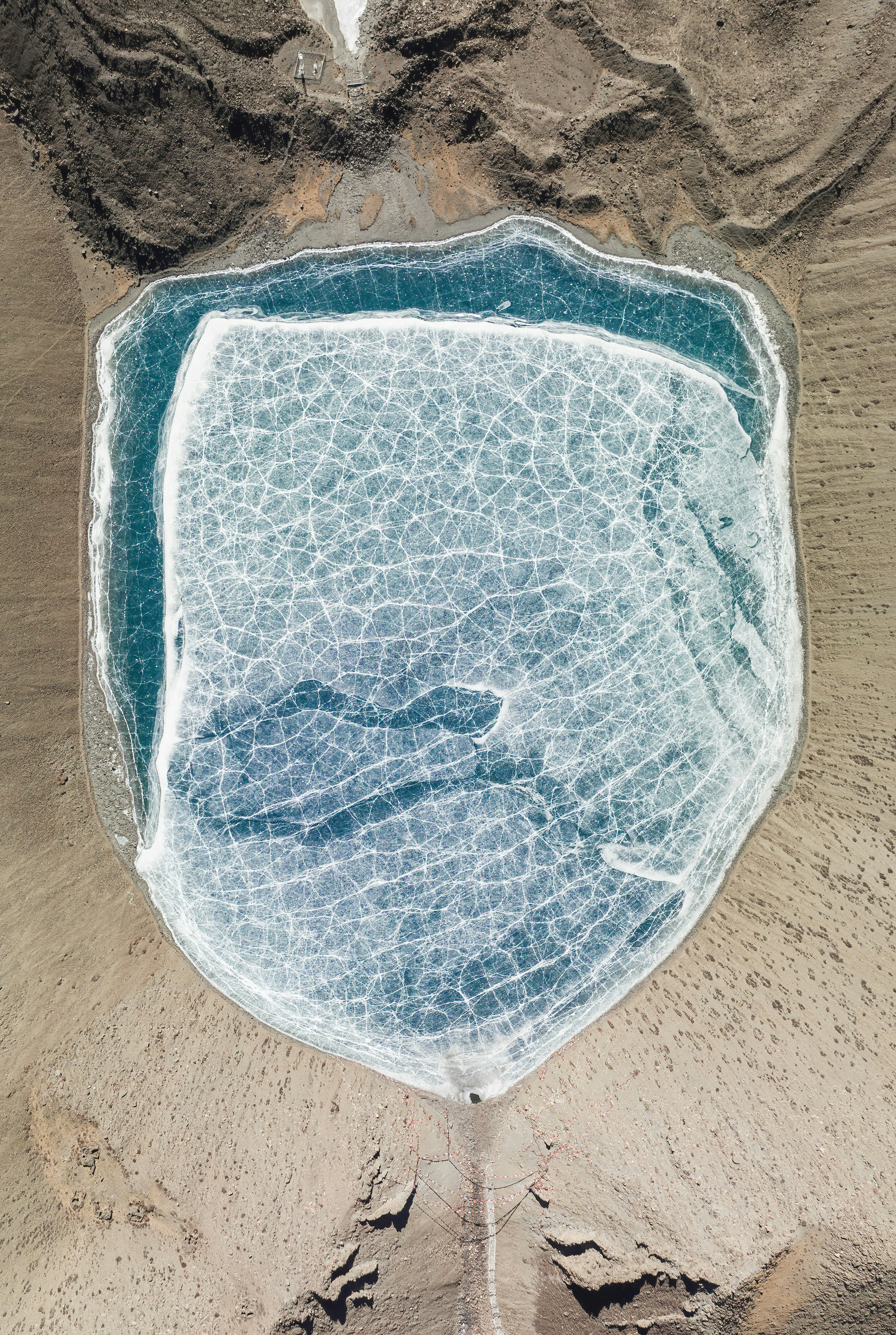 Aerial view of a frozen lake showcasing intricate ice patterns surrounded by barren terrain. The stark contrast highlights the beauty of winter's artistry.