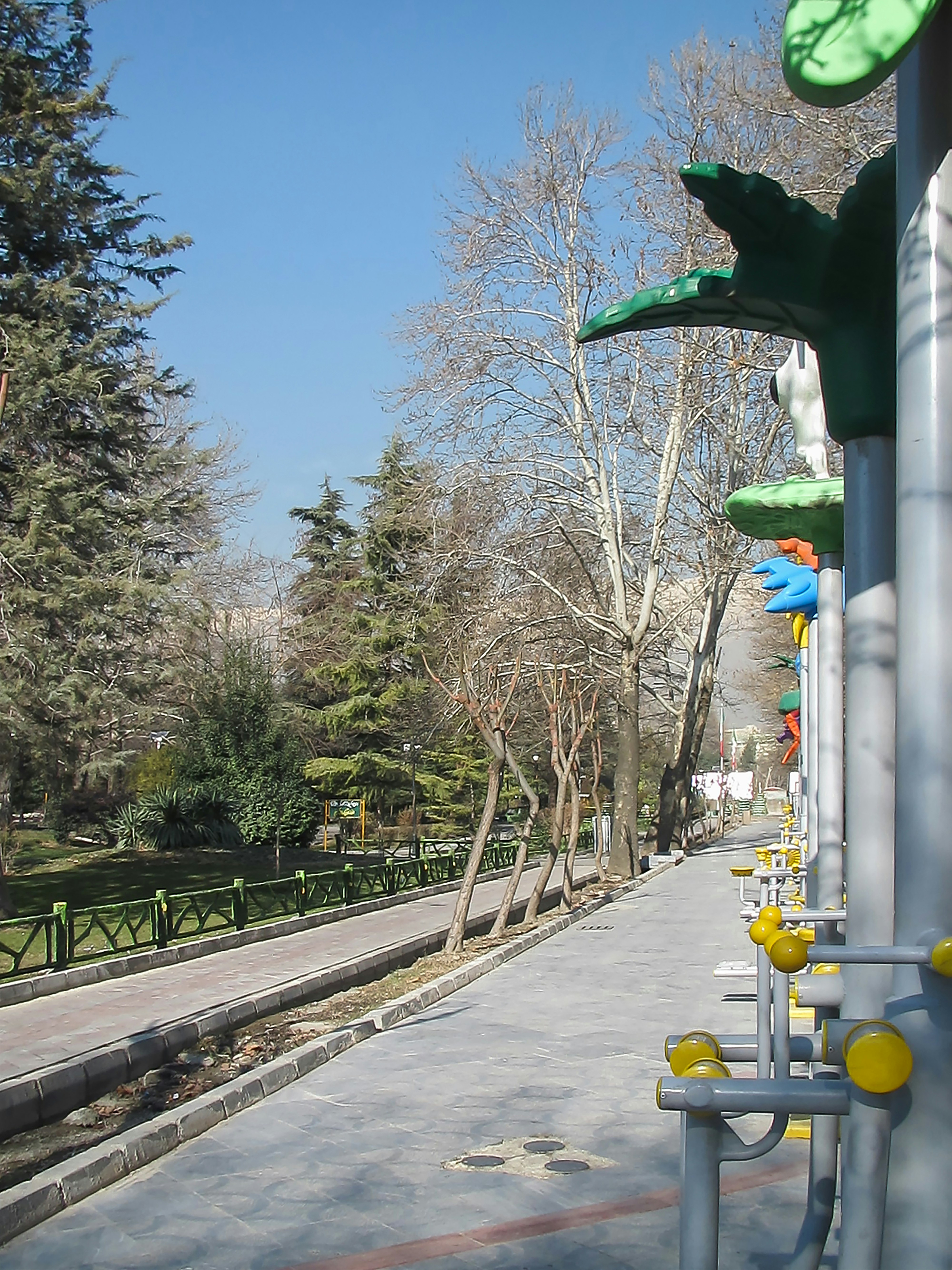 A sunlit park path flanked by trees on the left and a row of playful, multicolor hand-like sculptures along a railing on the right. The scene emphasizes depth and perspective as the walkway recedes into the distance.