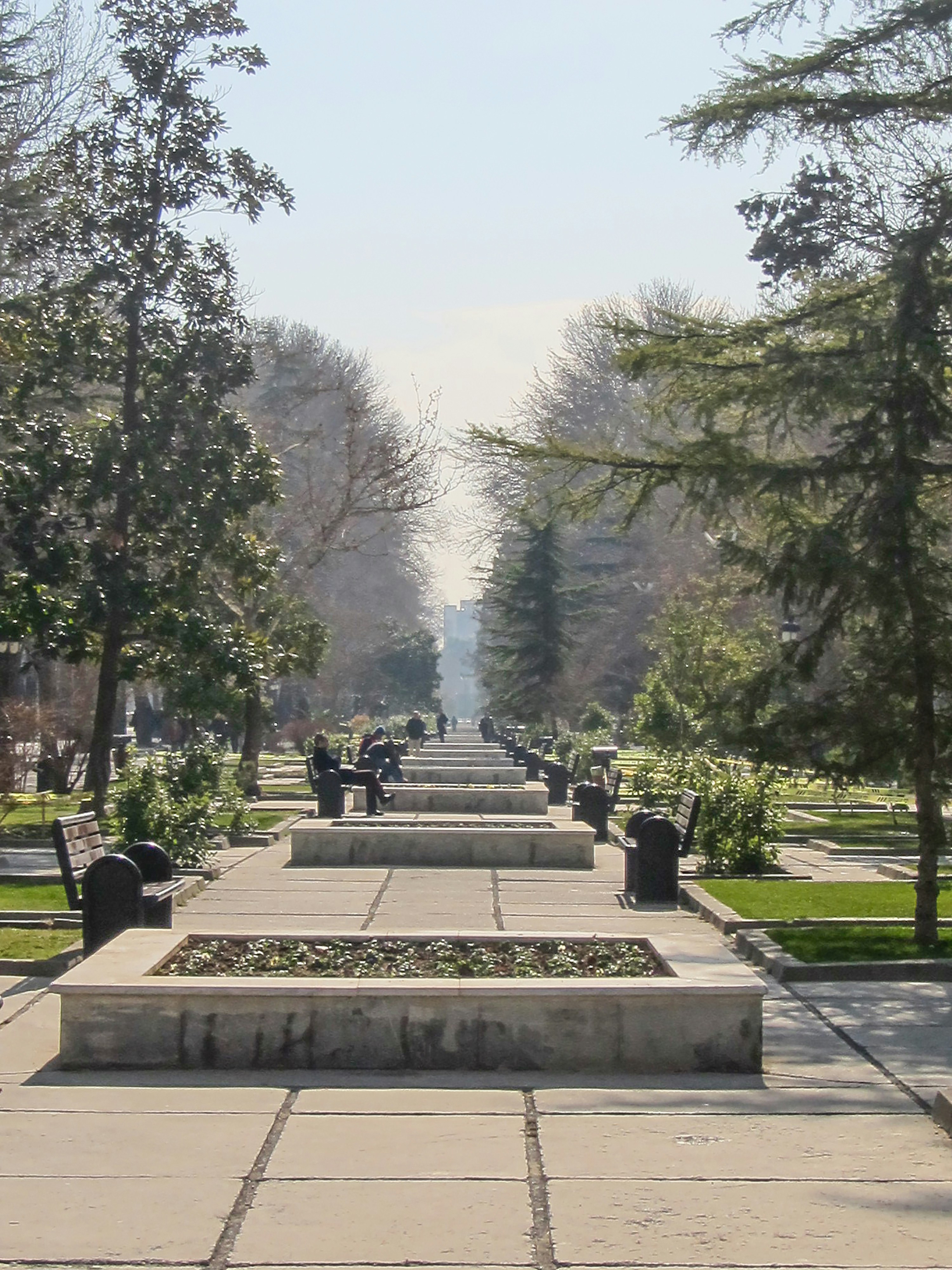 Wide park avenue with symmetrically placed planters and benches fading into the distance; a calm, sunlit perspective of a public garden.