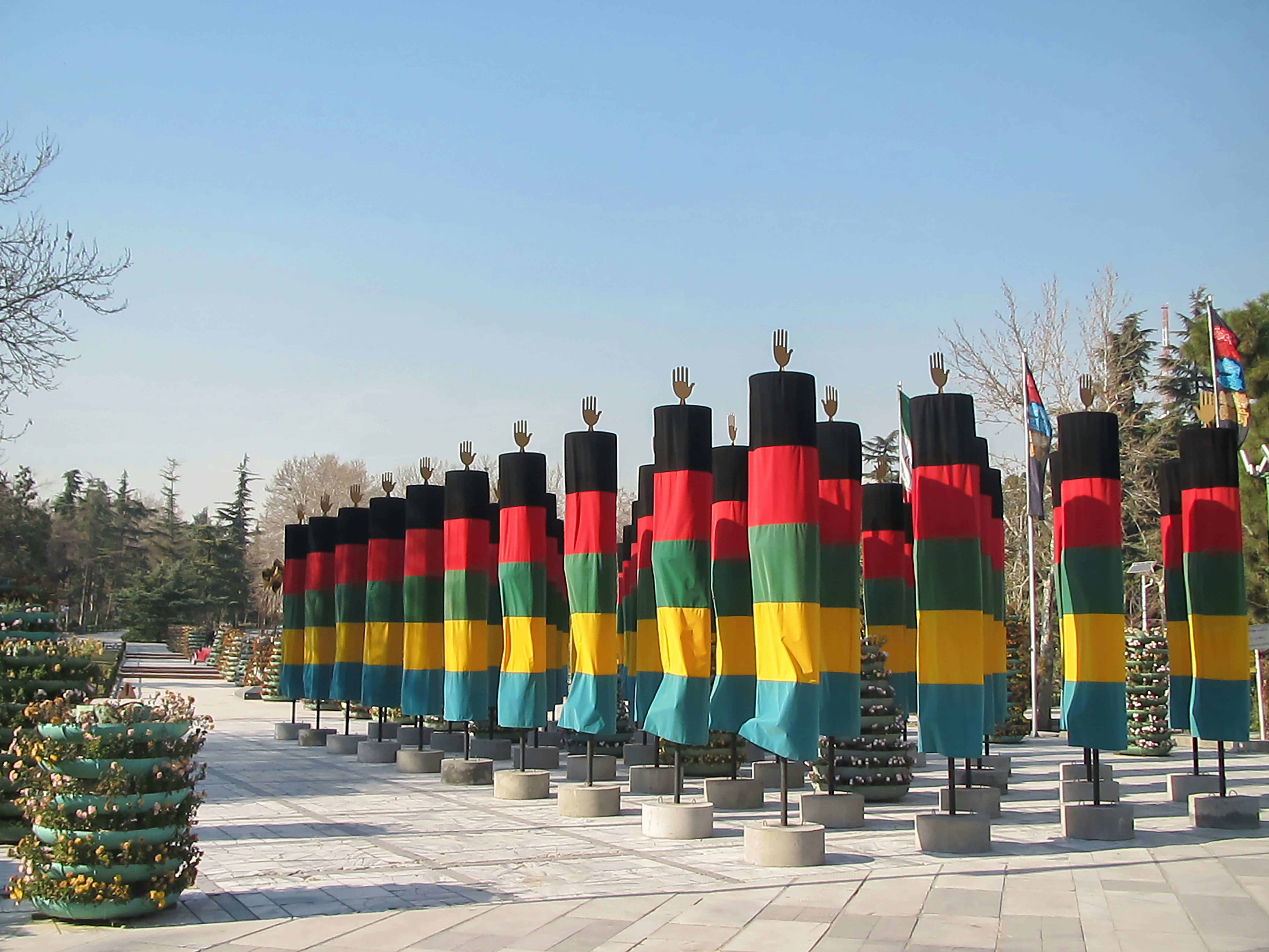Rows of tall vertical banners line a sunlit plaza, their bold color blocks (black, red, yellow, green) contrasting with the pale paving and clear sky.
