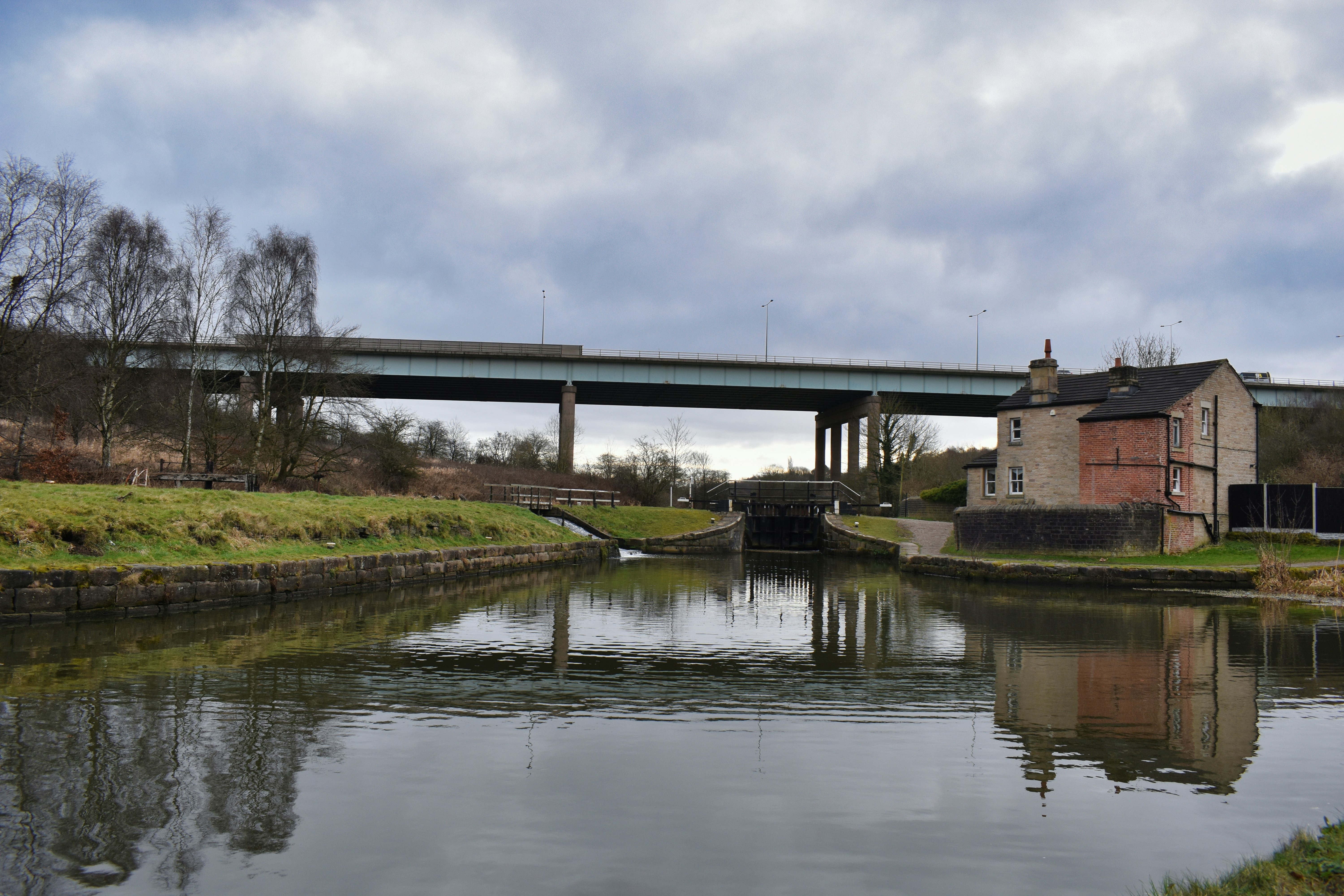A tranquil canal scene featuring a quaint house and a large bridge overhead, with reflections shimmering on the water's surface.