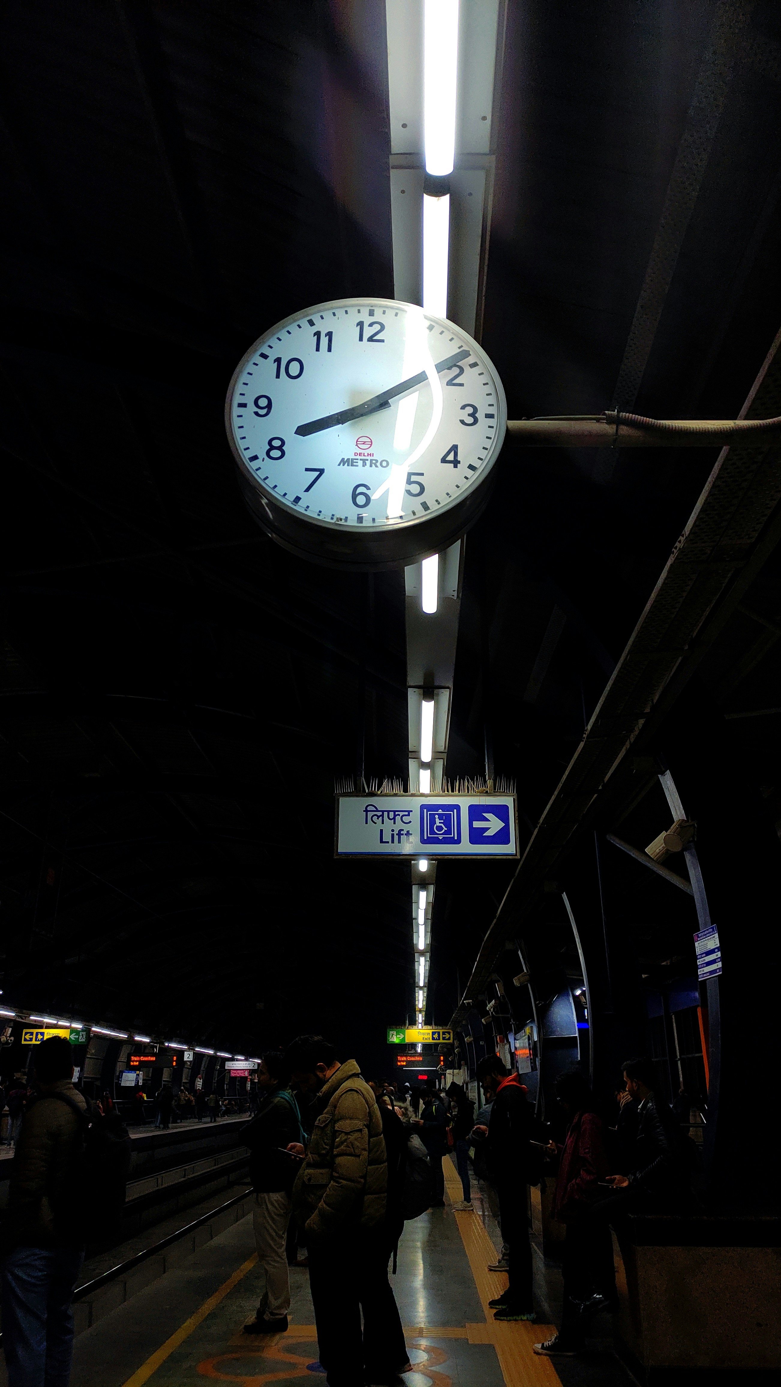 Clock displaying the time in a bustling metro station, with commuters waiting on the platform. Signage indicates lift access to different levels.