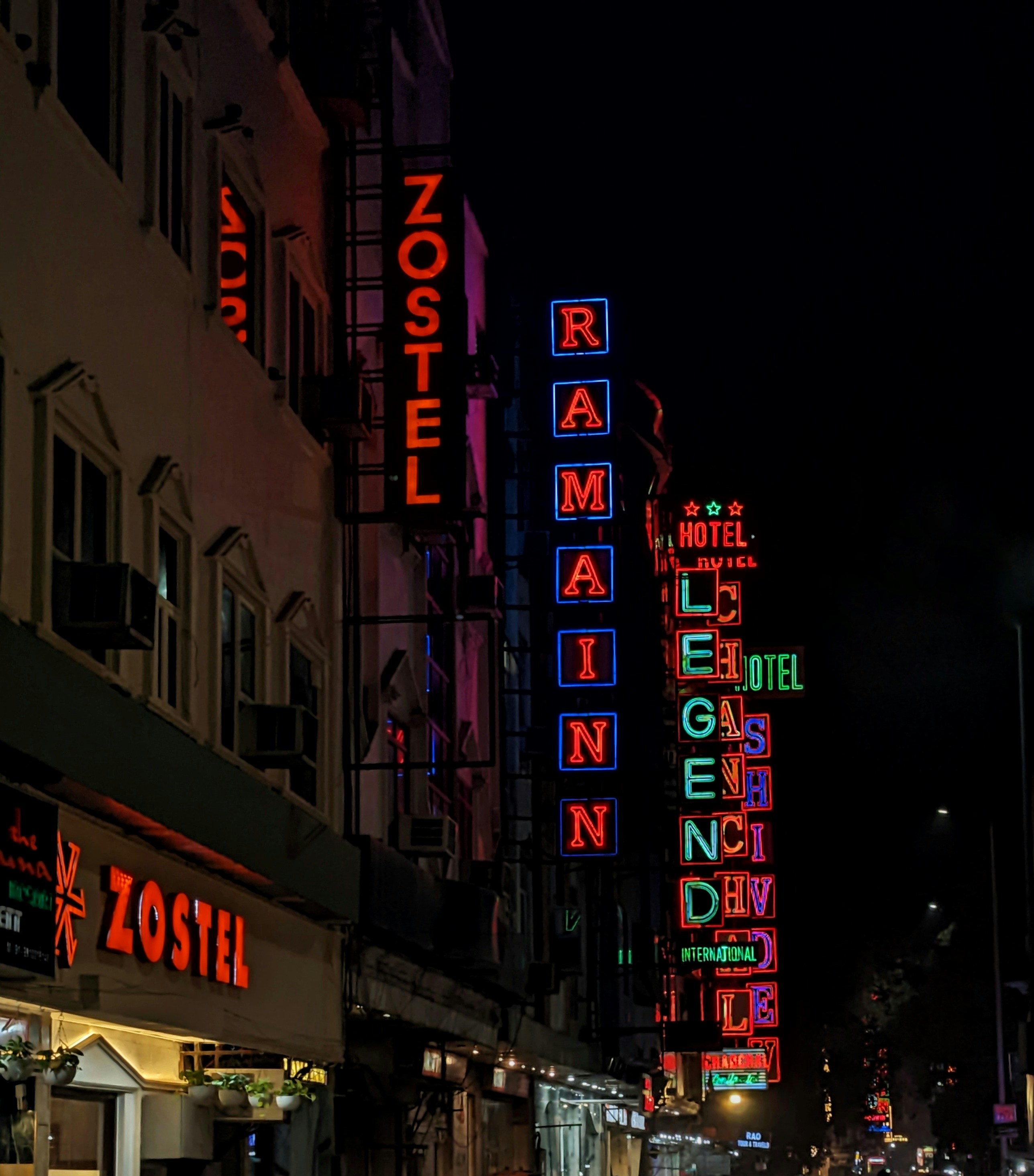 Bright neon signs illuminate the street, showcasing various hotels and hostels in a bustling urban setting at night.