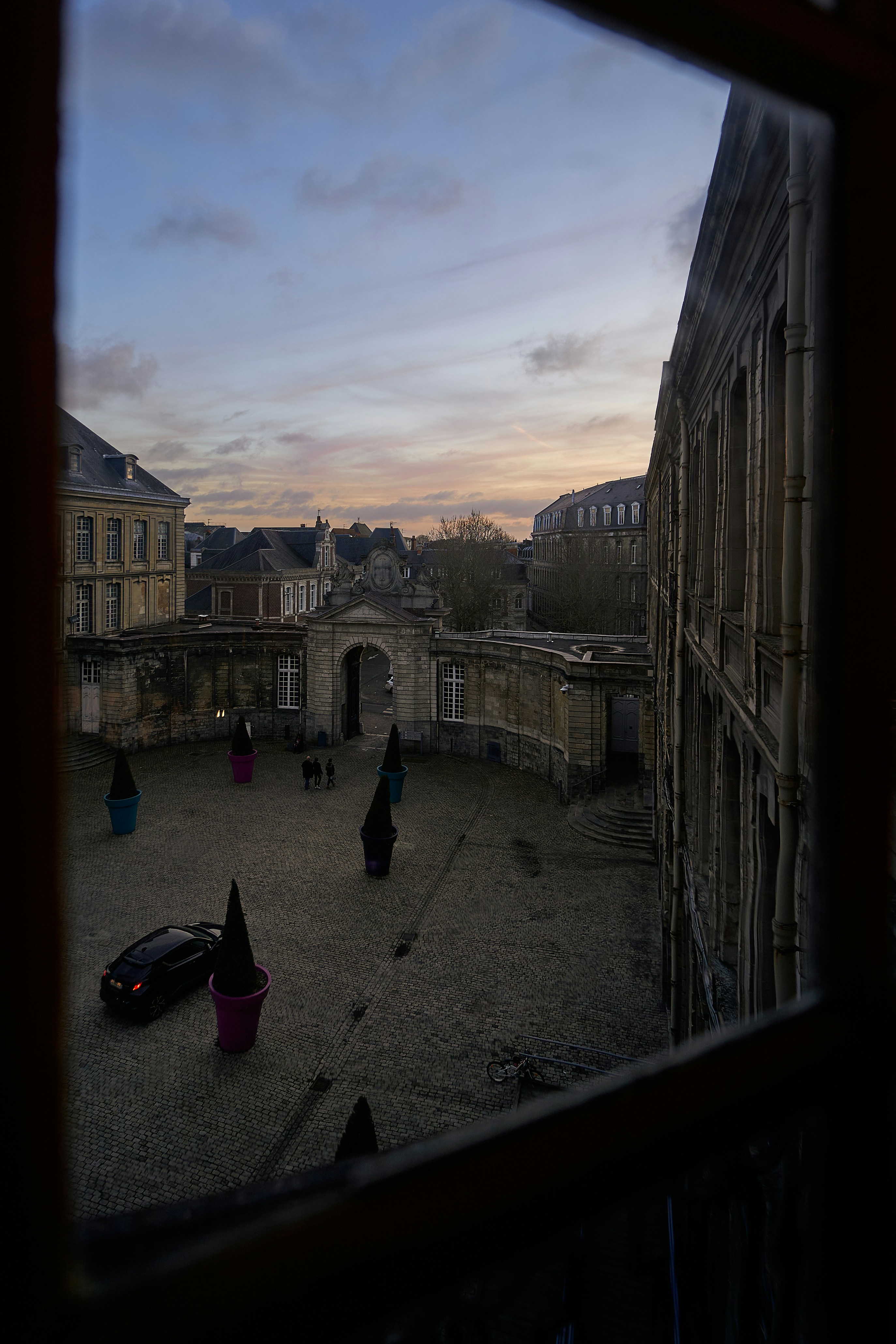 Courtyard view through a window at sunset, with historic architecture and a parked car.