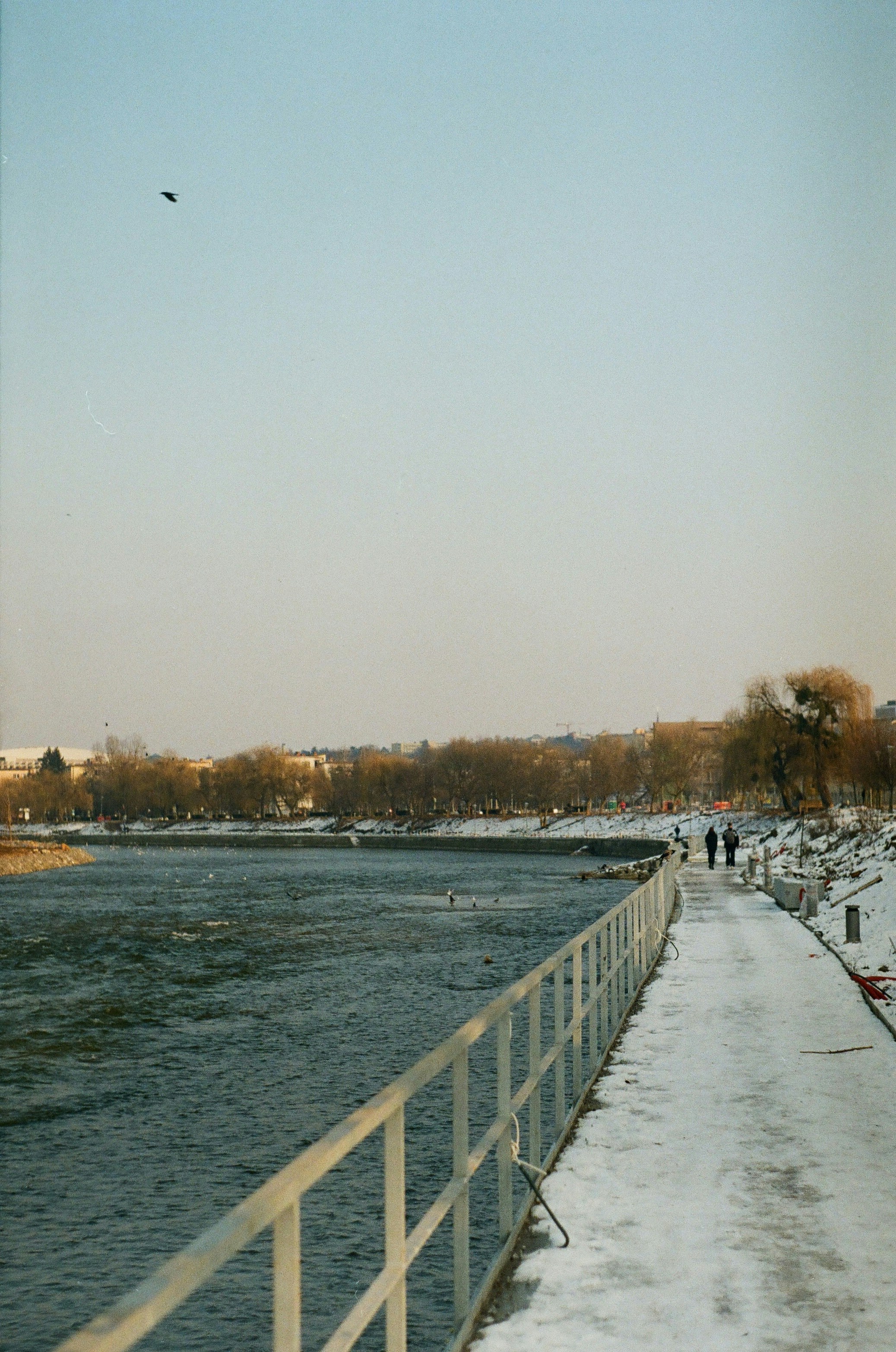 Frosted riverside walkway with a metal railing runs along a calm river, with distant walkers and snow-dusted trees under a pale winter sky.