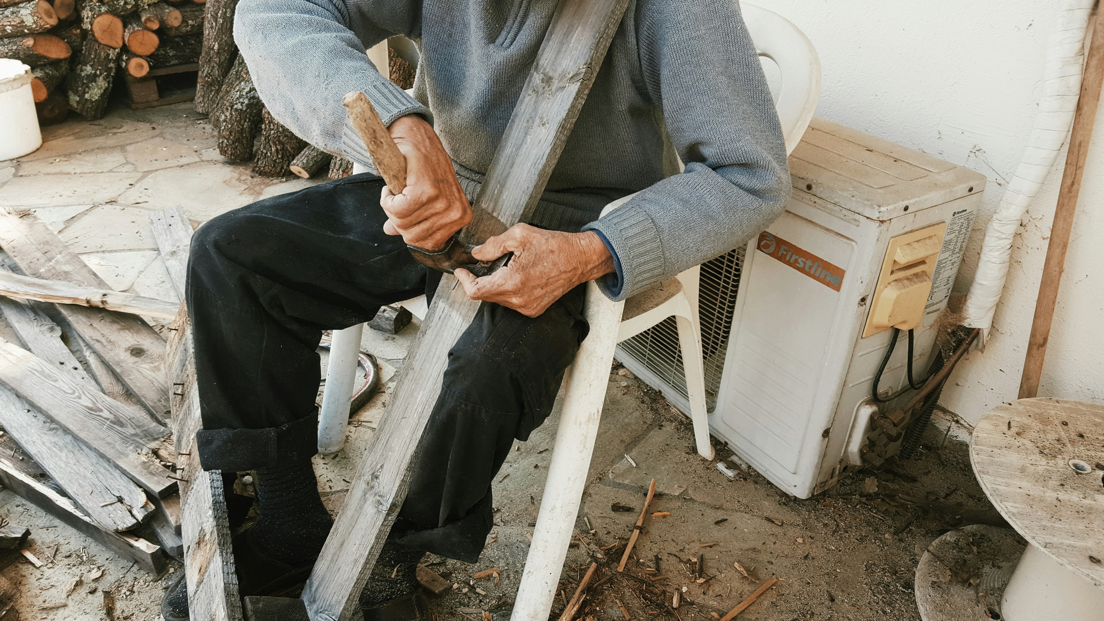 Skilled artisan meticulously carving wood while seated on a chair, surrounded by tools and materials in a rustic workshop.