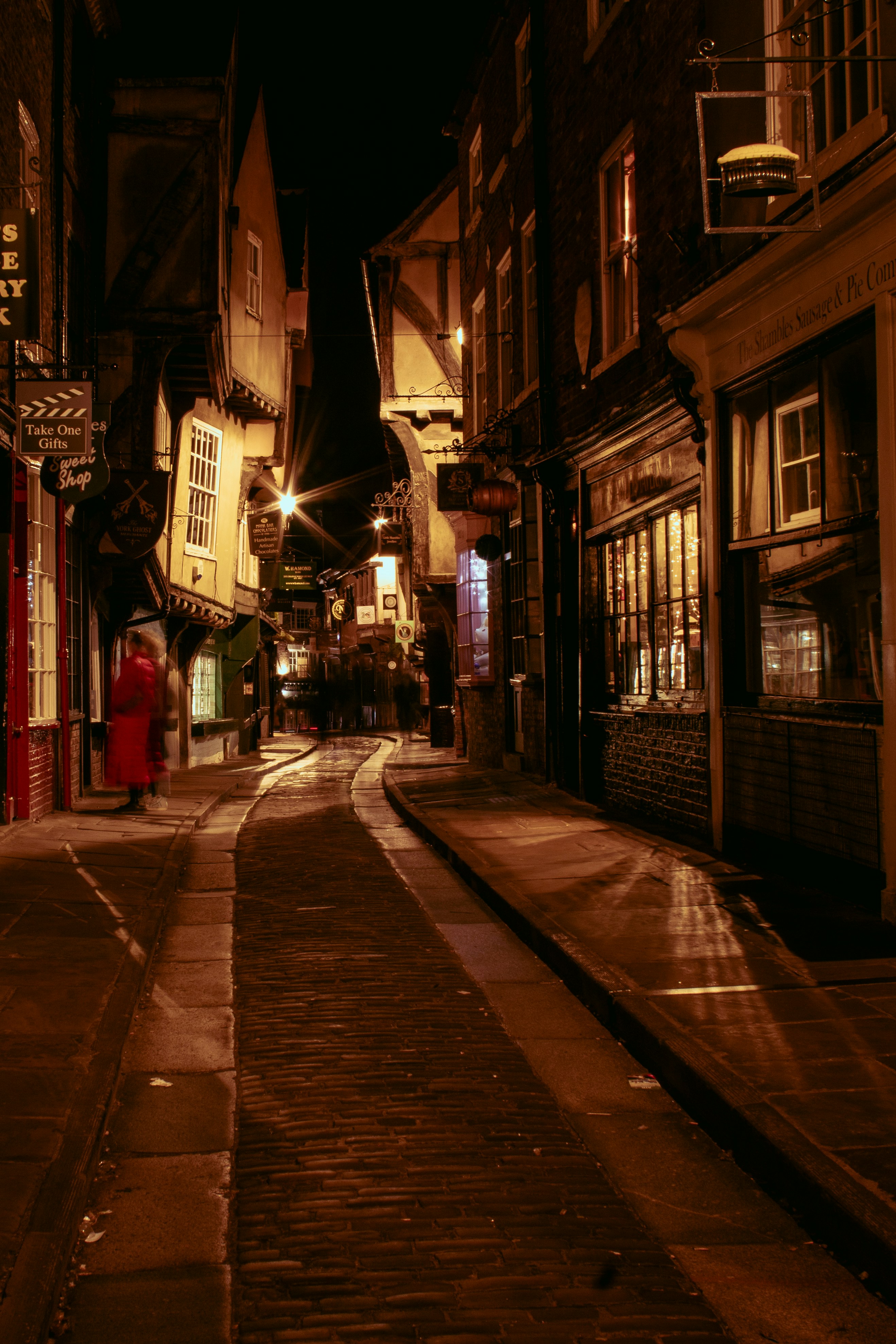 Narrow cobblestone street illuminated by warm streetlights, with a red fire hydrant and blurred pedestrian.