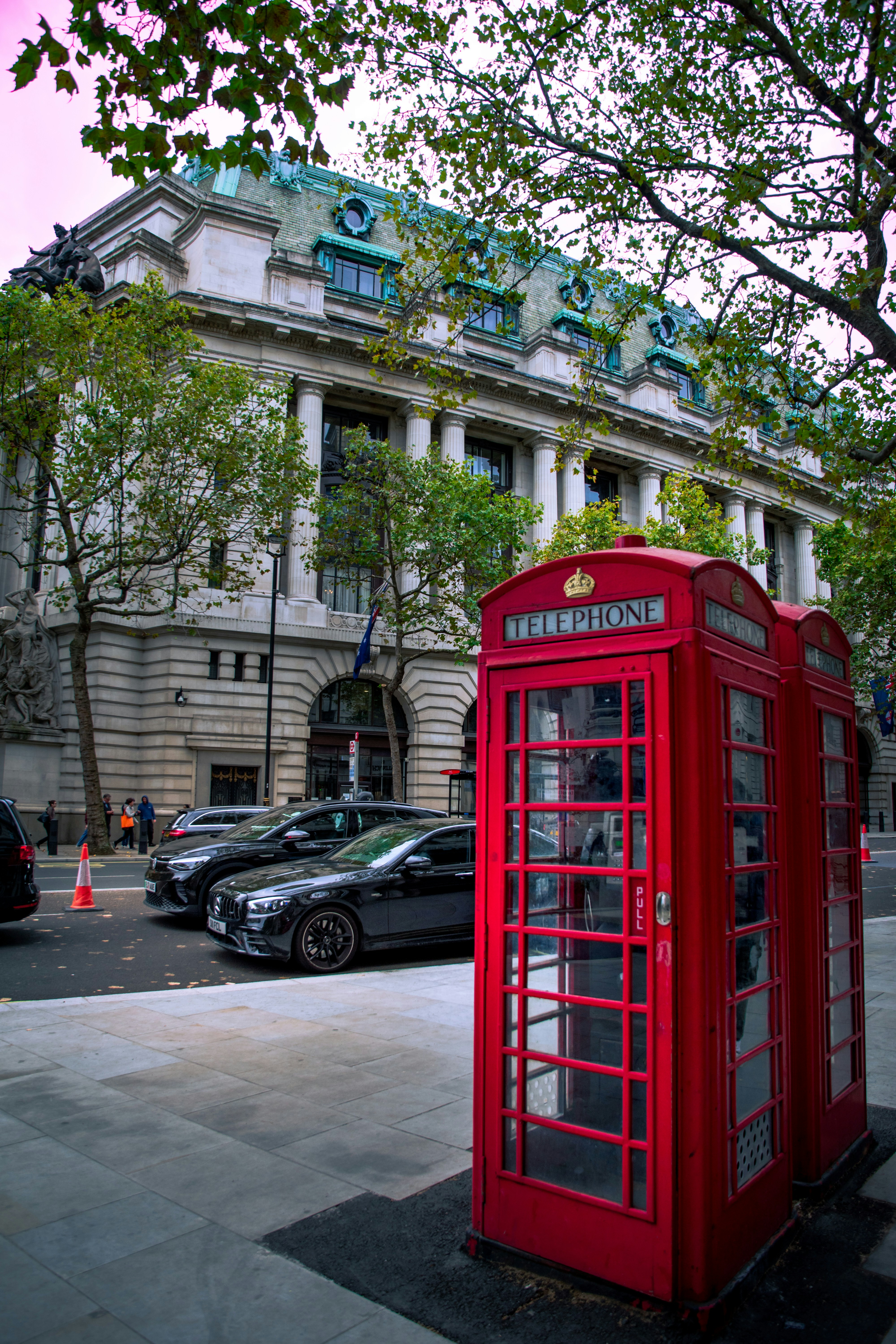 Two iconic red telephone boxes stand prominently on a bustling street, framed by an elegant historic building and lush trees.
