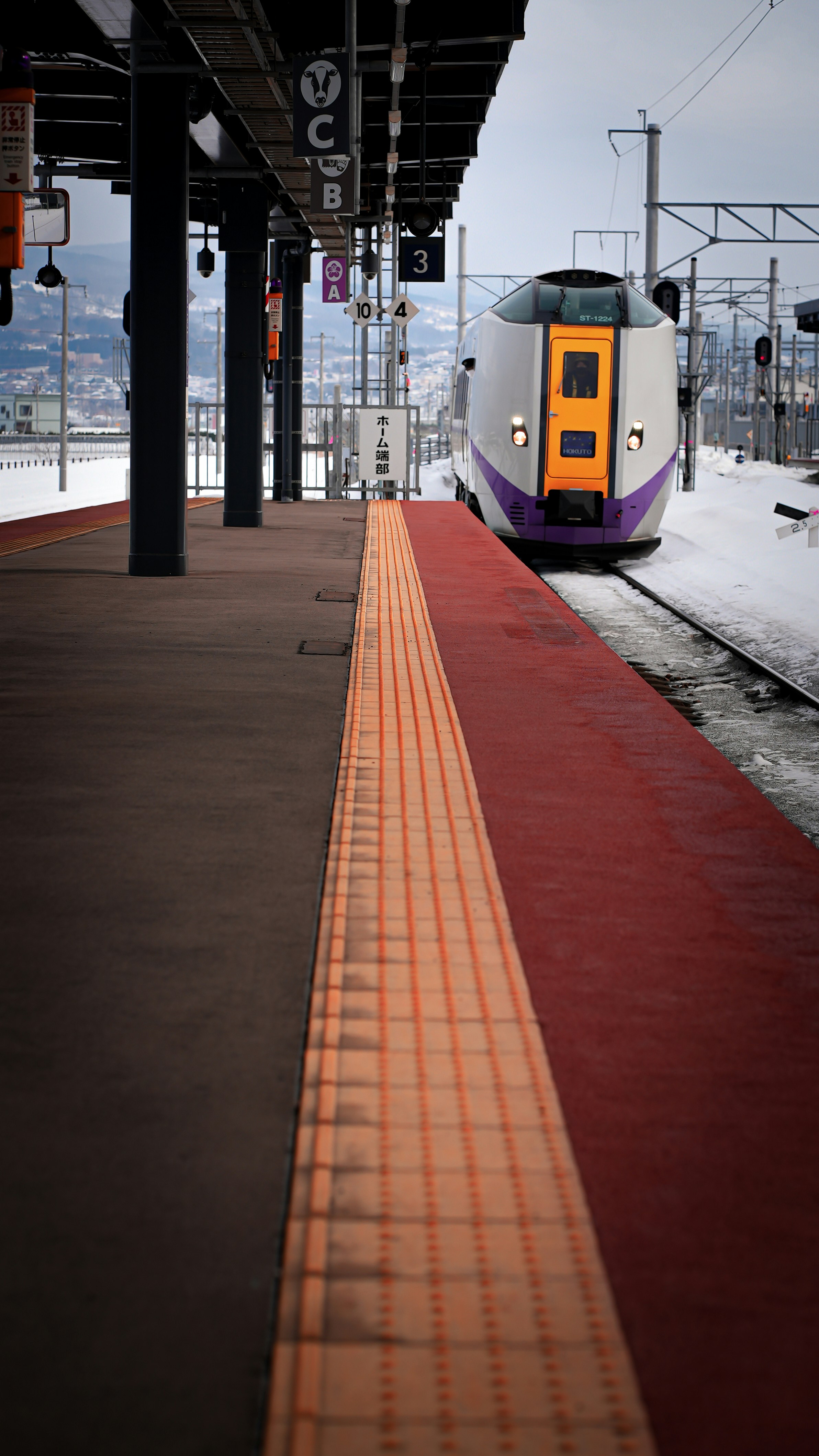 Train approaching a snowy station platform with vibrant orange tactile paving and signage in the background.