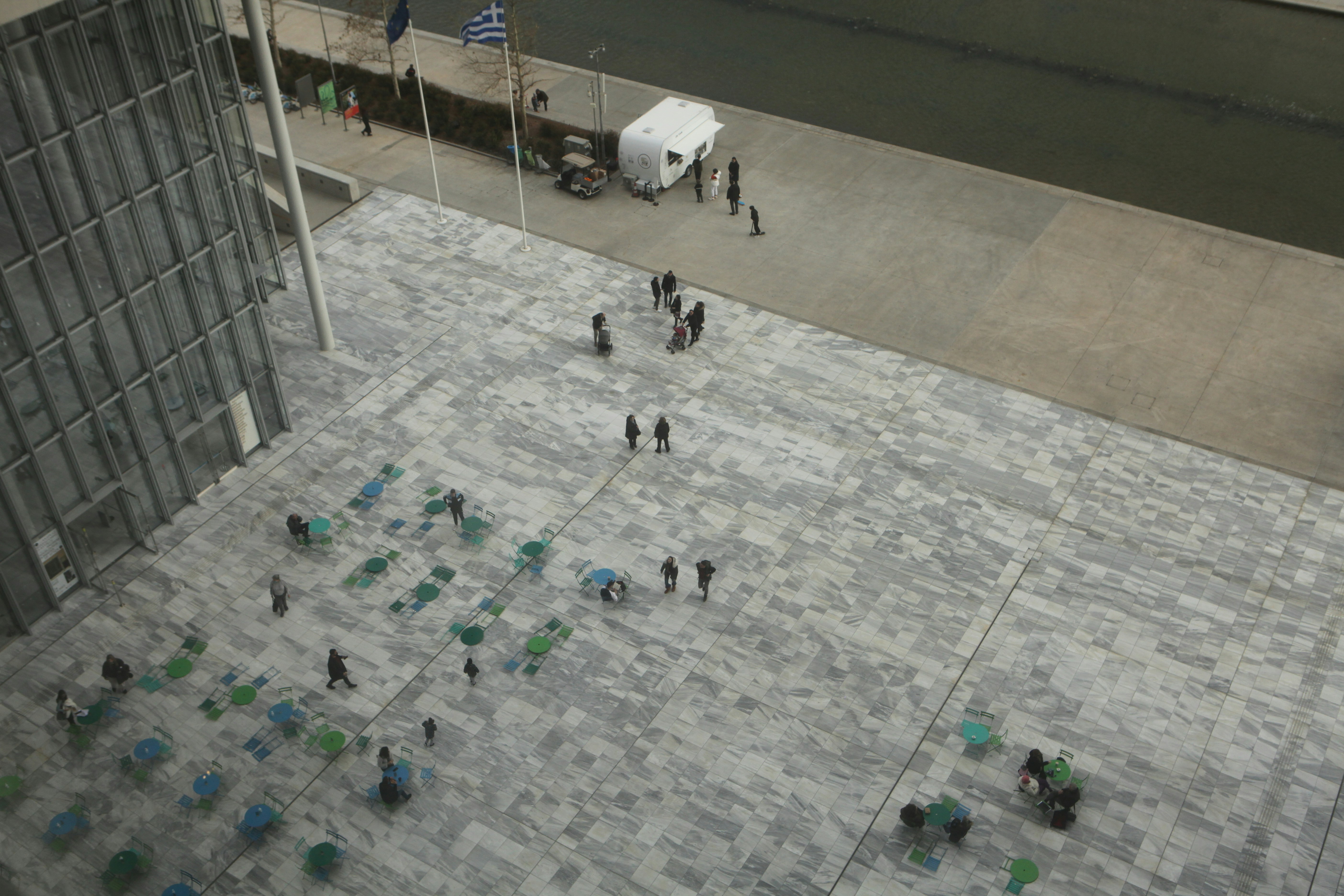 Aerial view of people walking across a large, tiled urban plaza with sparse green chairs.