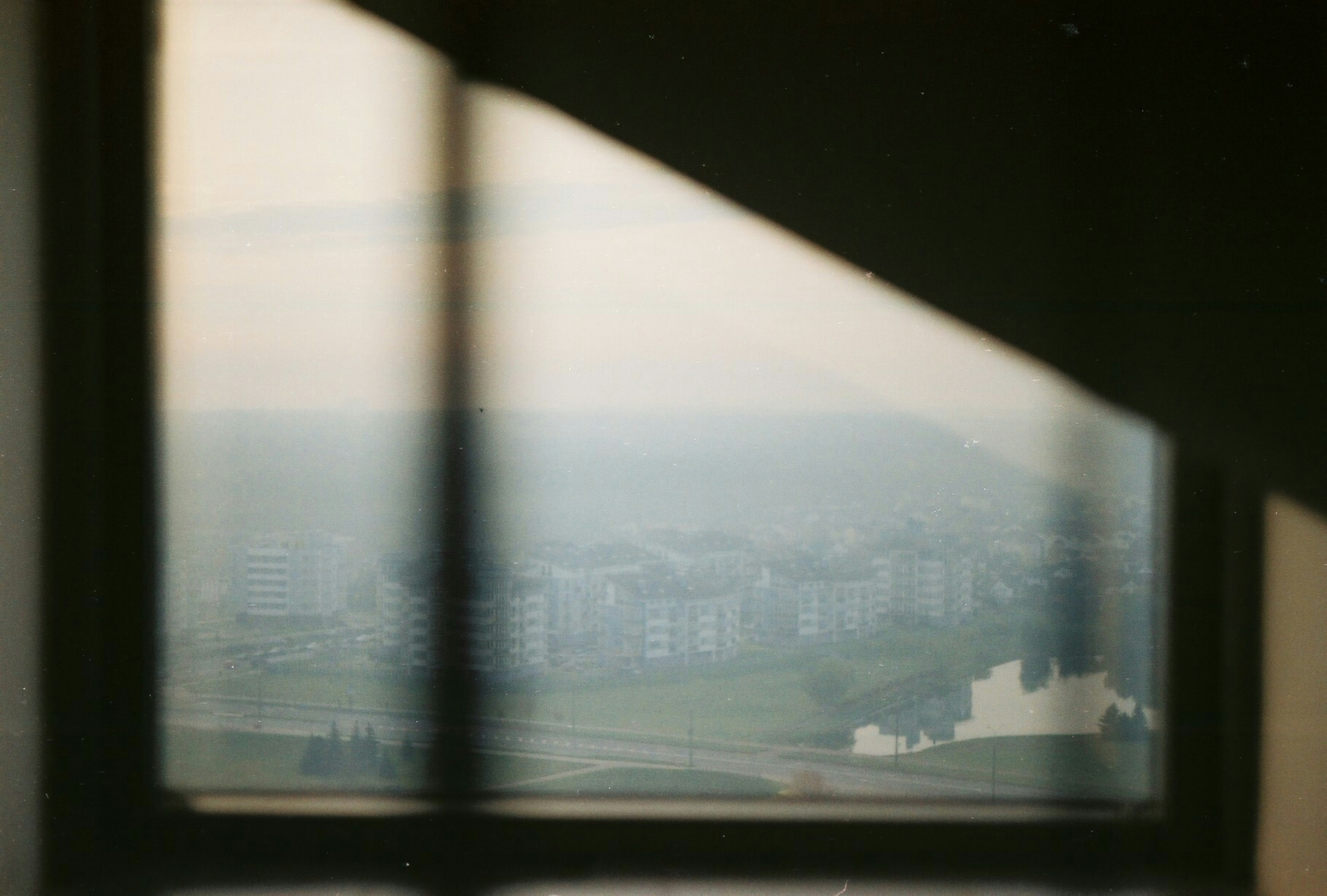 Soft, hazy photograph viewed through a window frame reveals a canal and low-rise apartment blocks in a muted urban landscape. Reflections and vertical window lines add depth and abstraction to the scene.