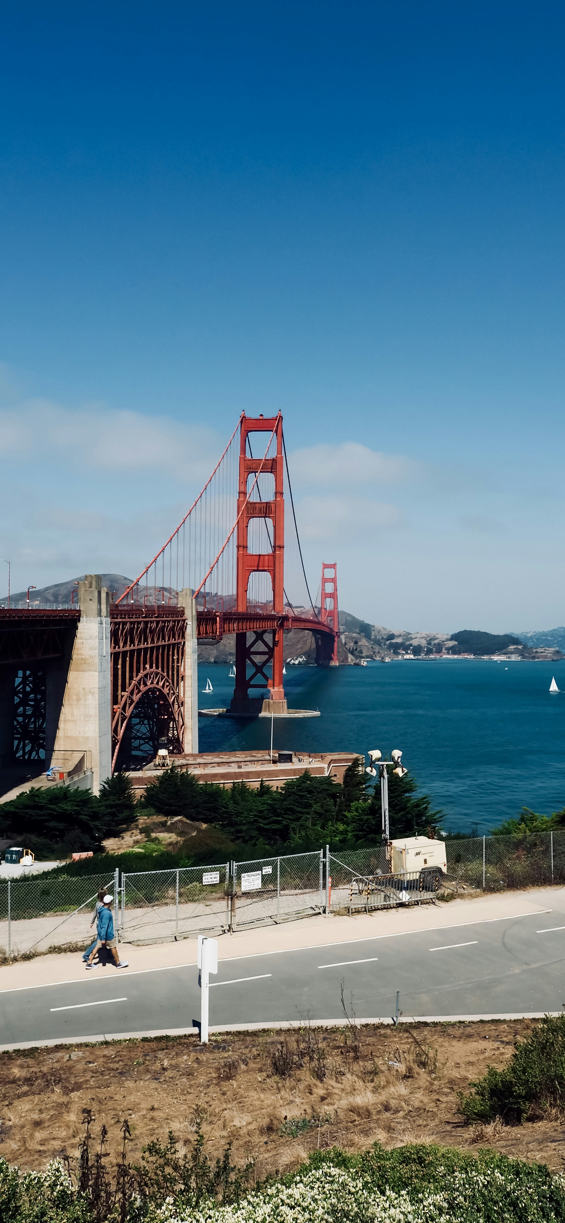 A photograph of the Golden Gate Bridge's red towers rising over the blue bay, with a fenced walkway and a pedestrian in the foreground.