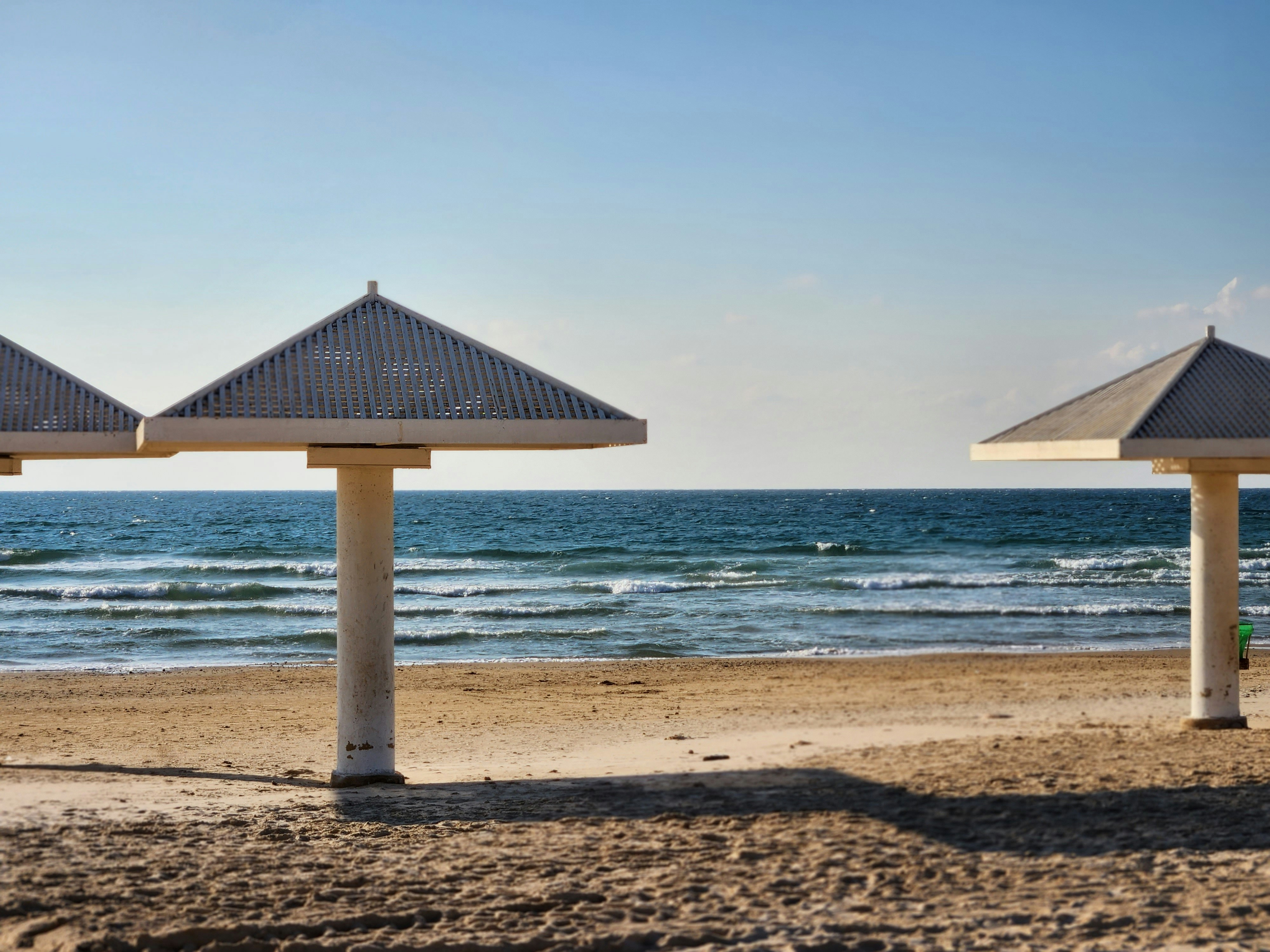 Three geometric parasols line a sandy beach with gentle waves under a clear sky.