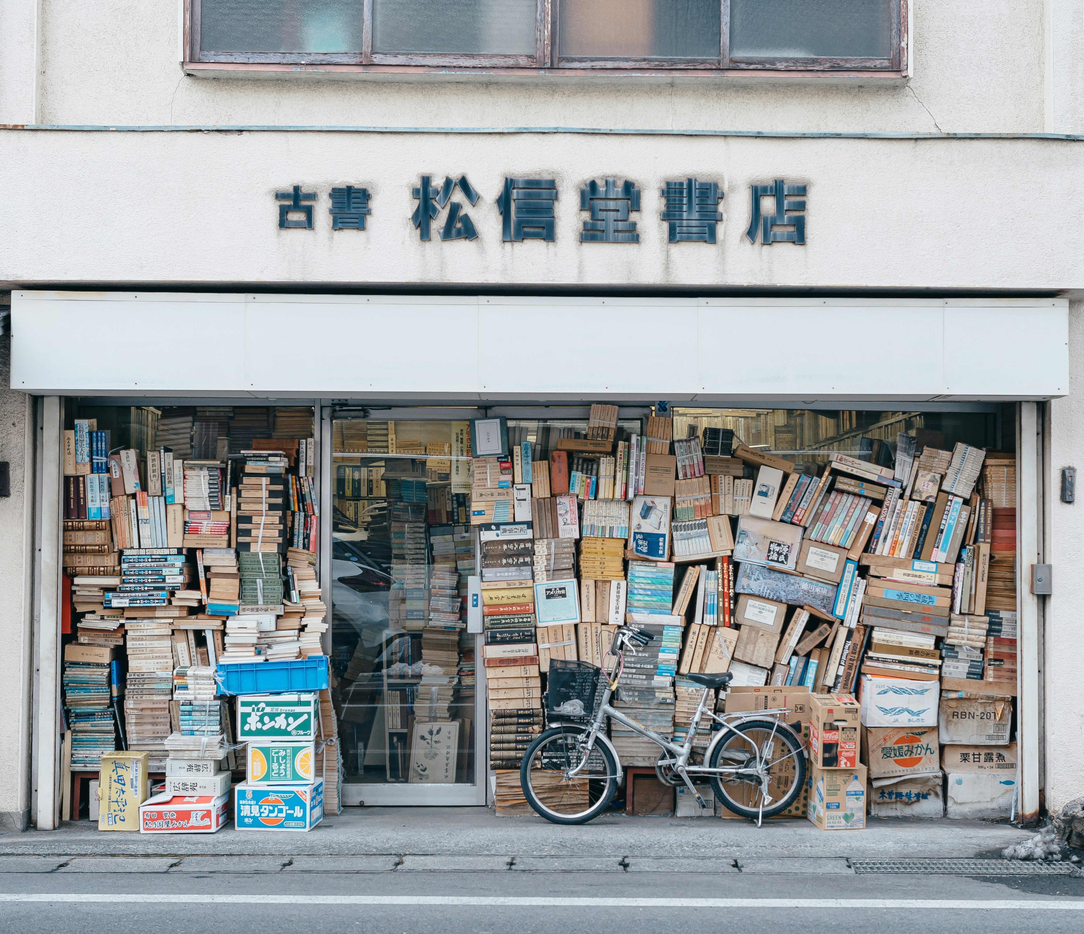 A quaint bookstore front overflowing with stacked boxes of books, accompanied by a parked bicycle. The shop's sign hints at its literary treasures.
