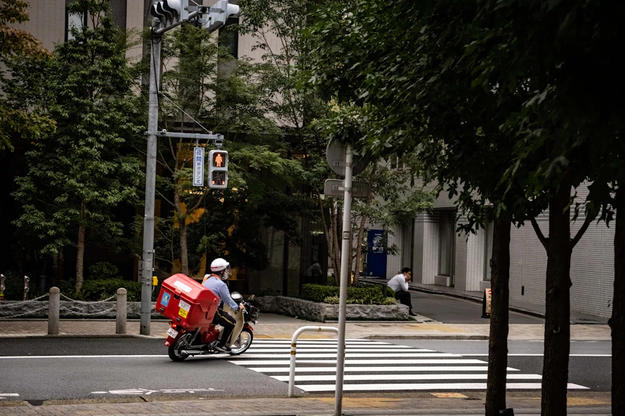 A red delivery scooter with a rider in a white helmet crosses a striped crosswalk on a tree-lined urban street. A pedestrian signal glows above the intersection, with buildings and foliage in the background.