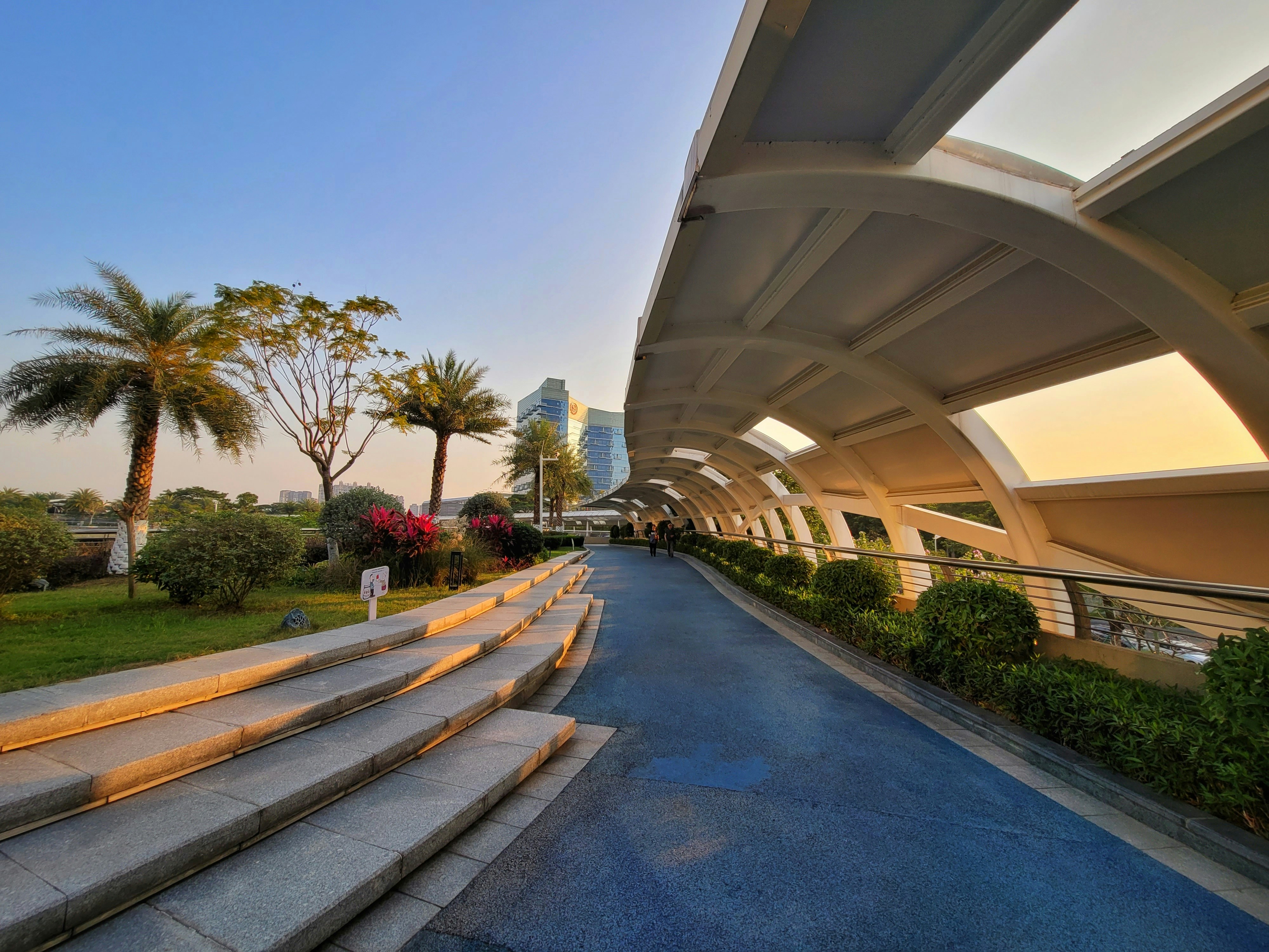 Curved walkway flanked by lush greenery and futuristic architecture under a clear sky.