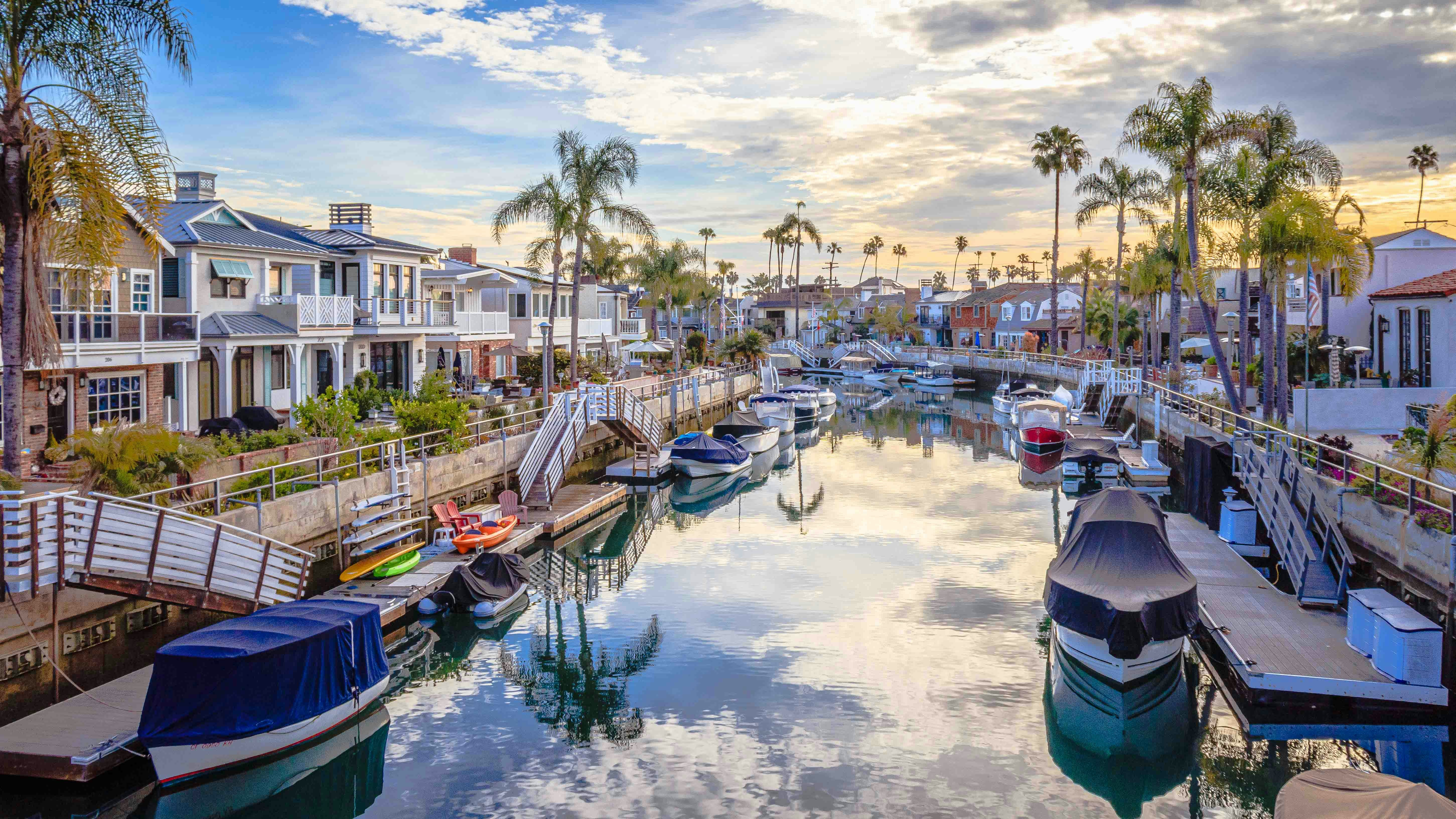 Boats docked along a serene canal lined with palm trees and houses under a partly cloudy sky.
