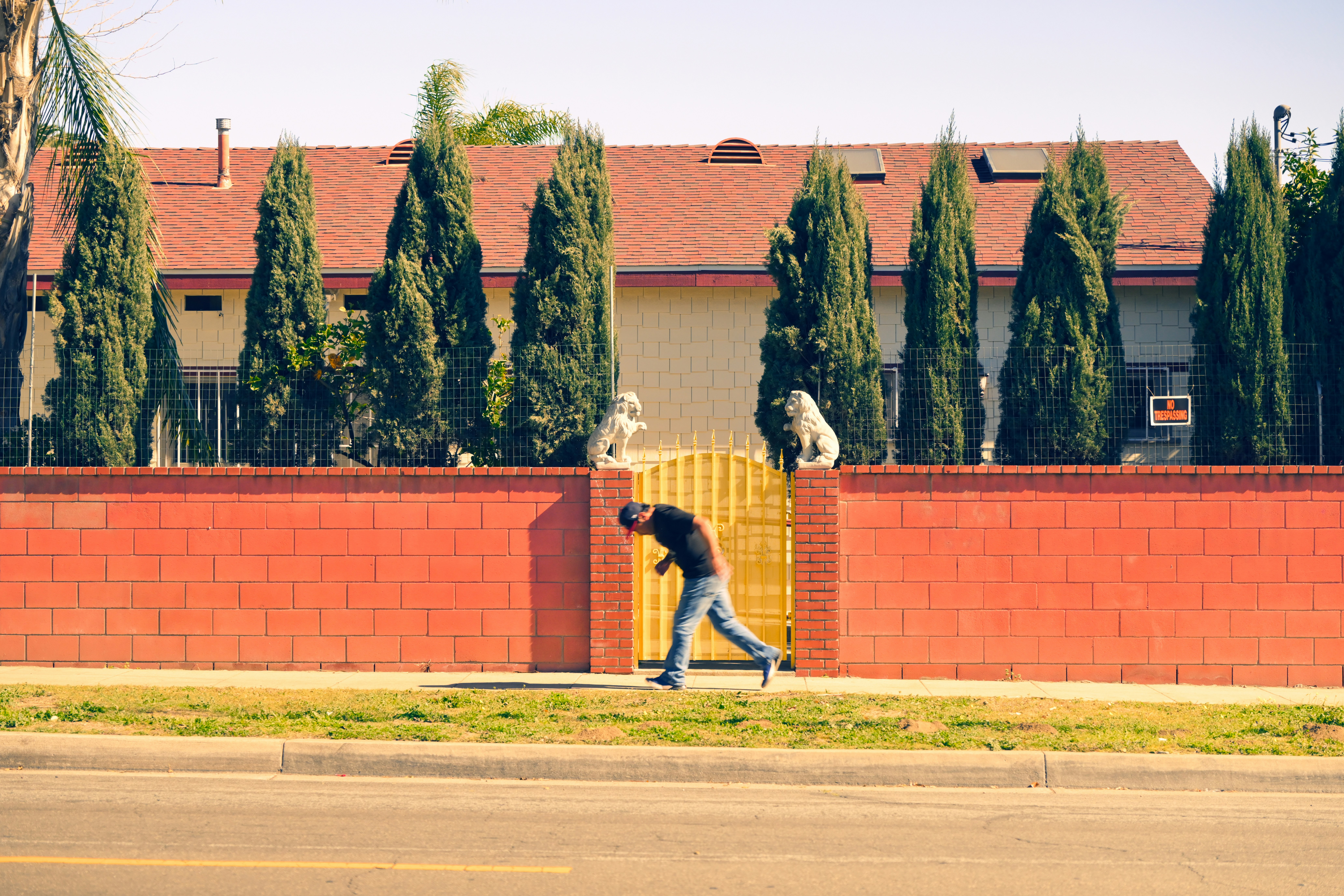 Man walking along a red brick wall adorned with statues and tall trees, leading to a gated entrance. The scene captures a blend of urban life and decorative elements.