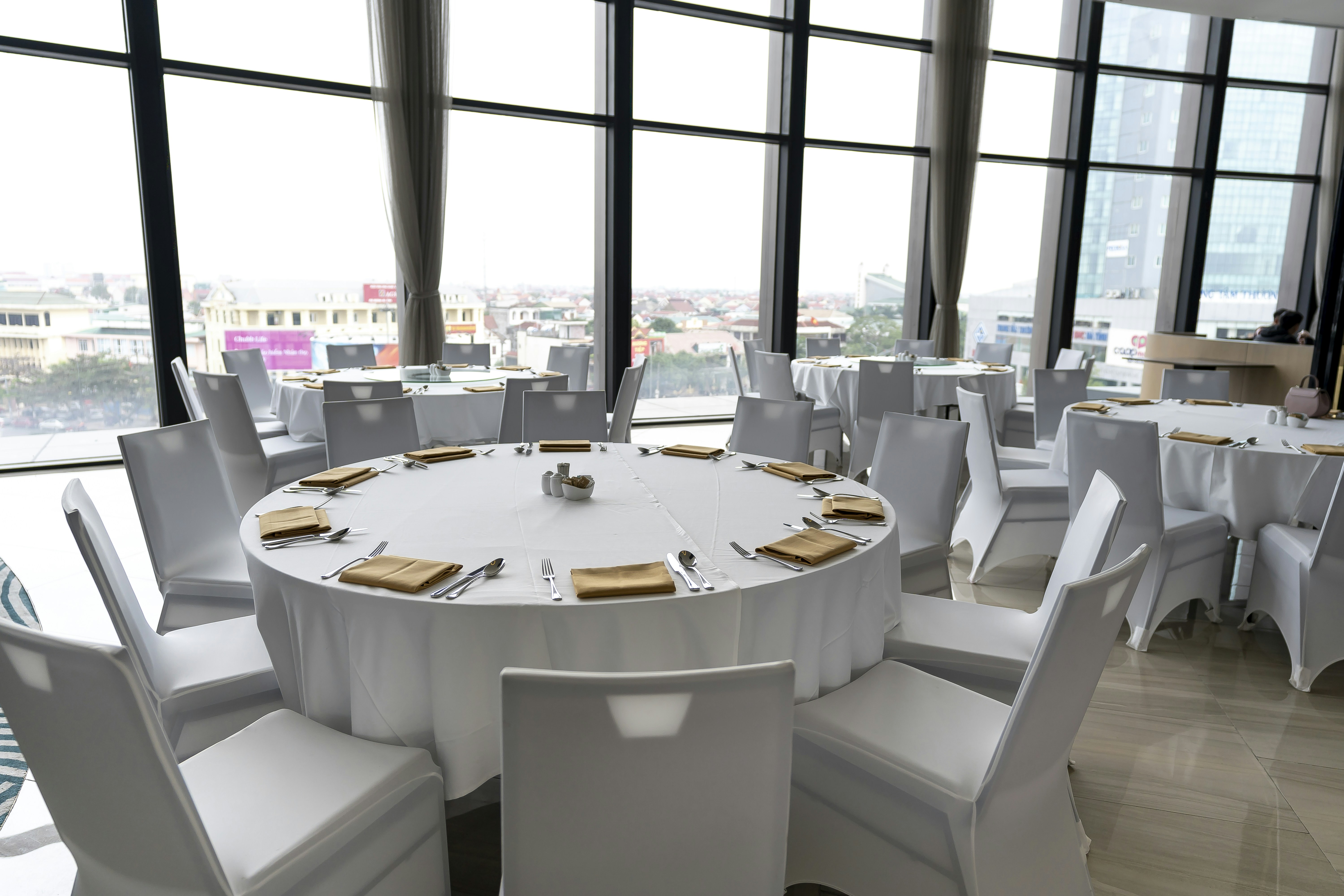 Contemporary dining setup featuring a round table adorned with neatly arranged cutlery and golden napkins, set against a backdrop of large windows revealing a cityscape.