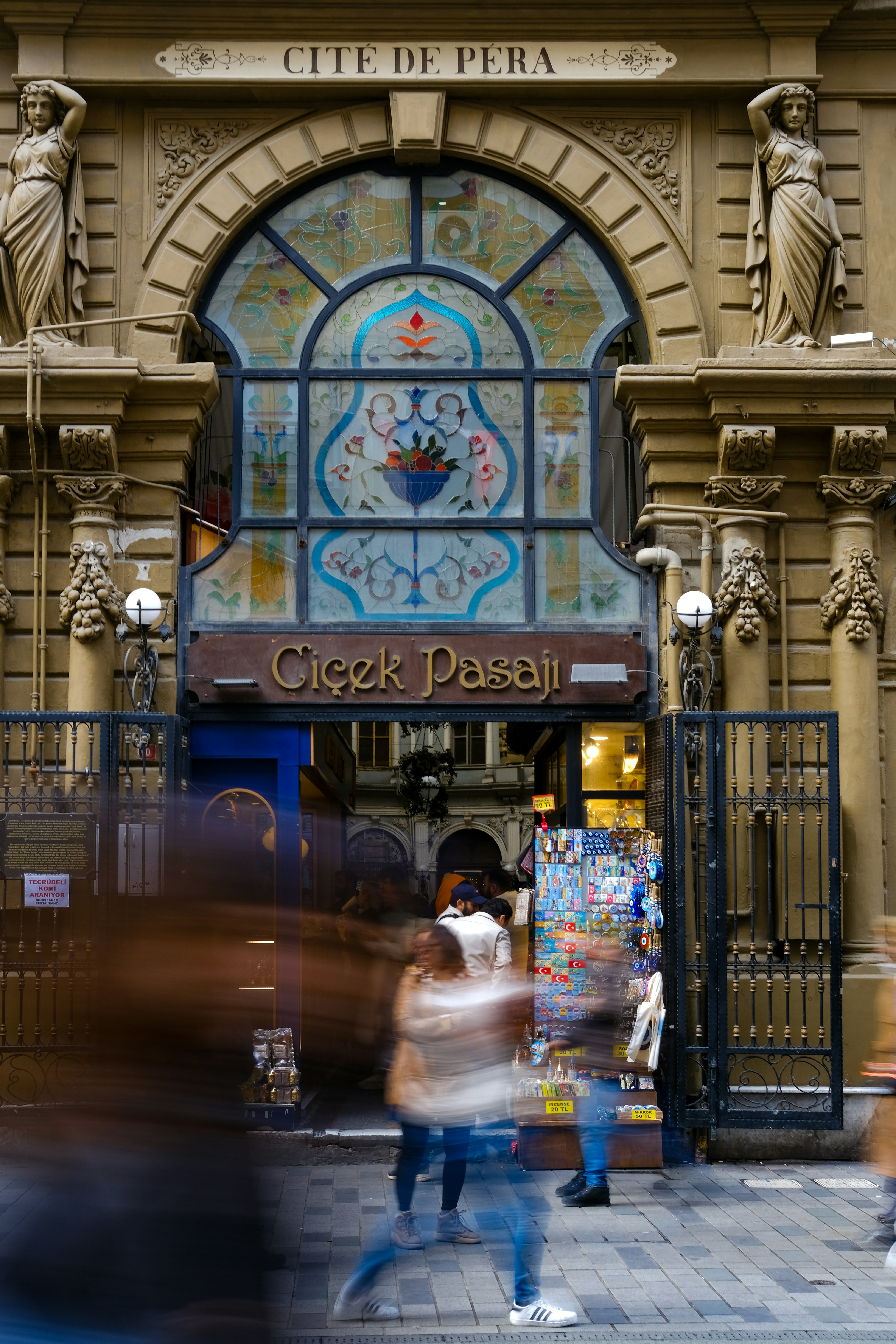Art nouveau entrance of Cicek Pasaji adorned with stained glass, bustling with pedestrians and colorful merchandise on display.