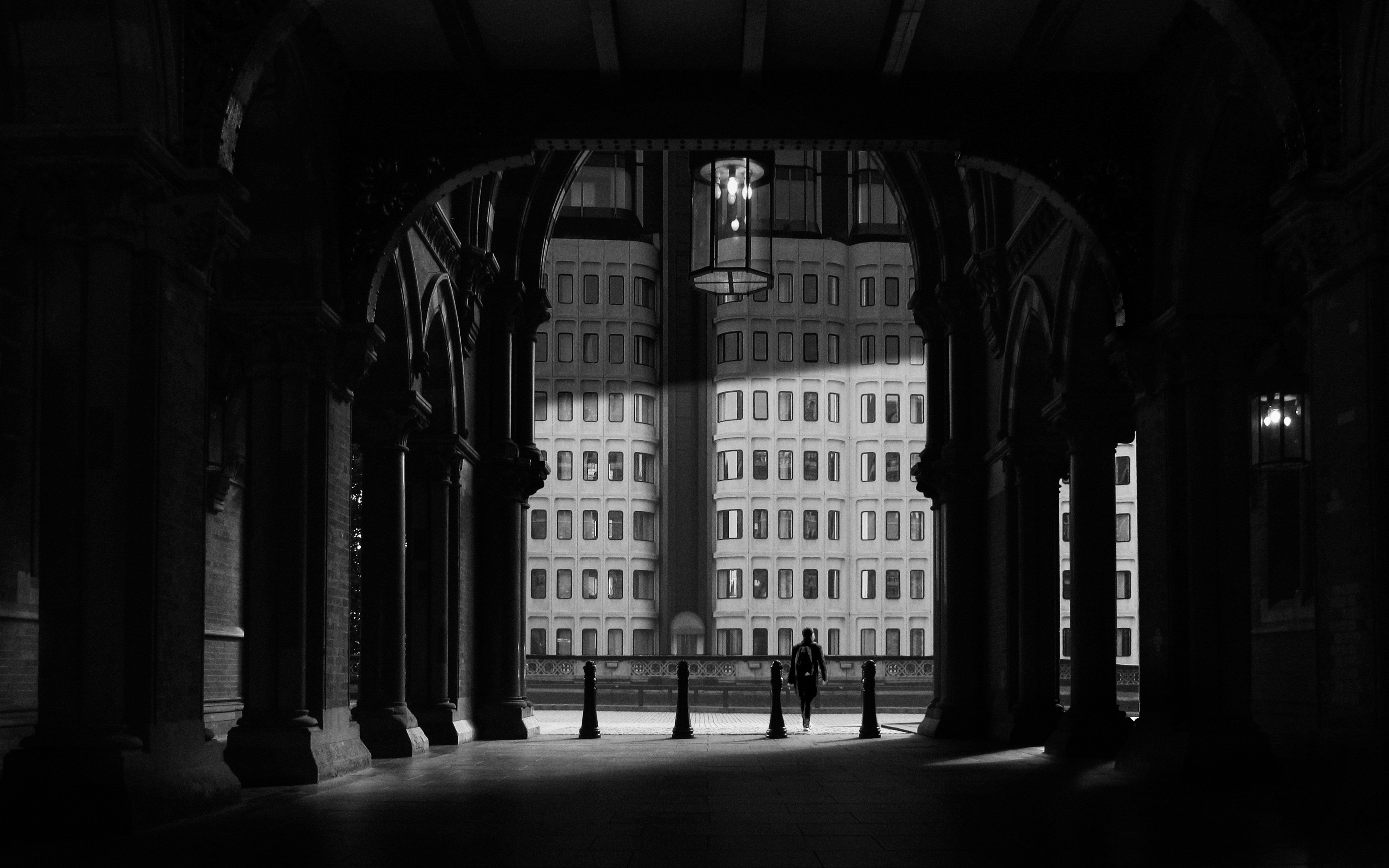 Silhouetted figures stand beneath an archway, framed by contrasting light and shadow, revealing a glimpse of an architectural backdrop.