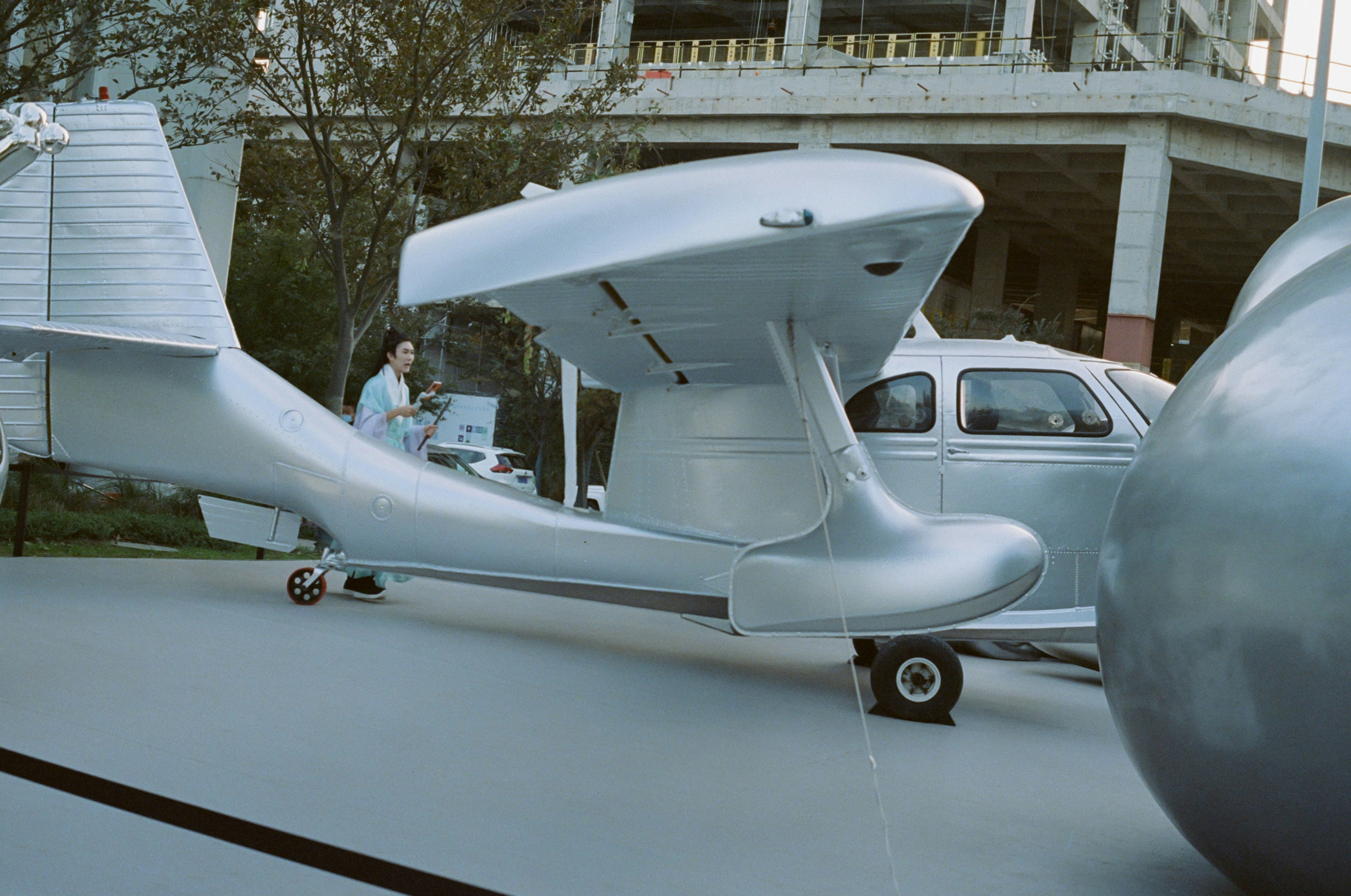 A sleek silver aircraft stands beside a vintage car, showcasing a blend of modern and classic automotive design in an urban setting.