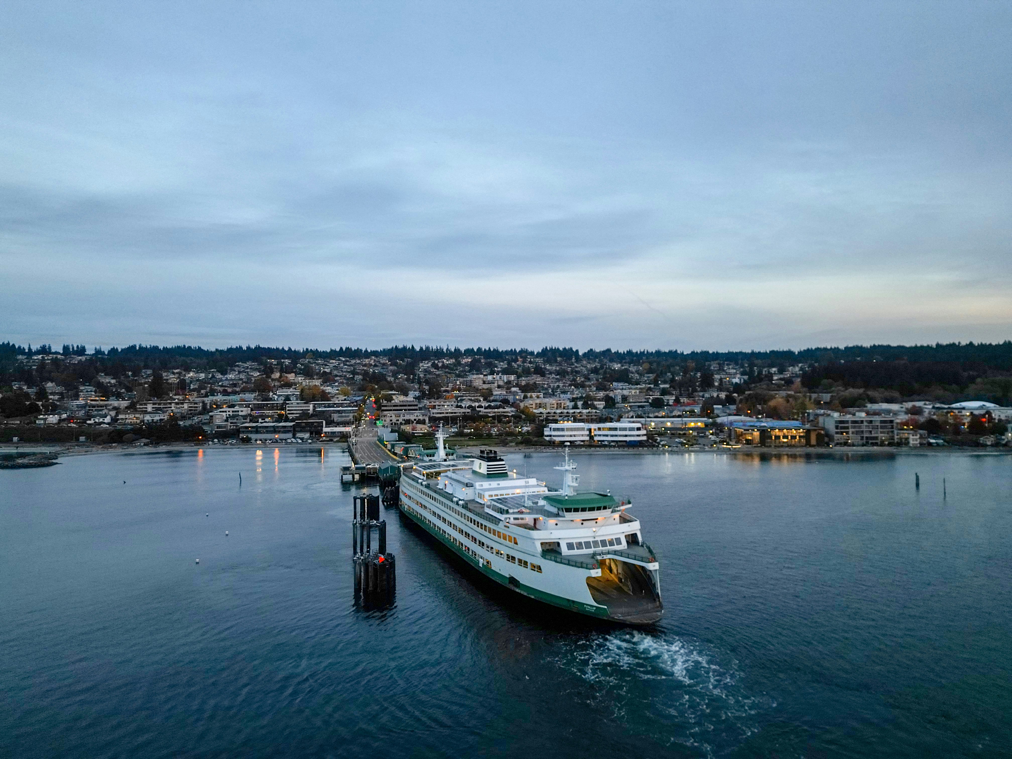 Ferry navigating calm waters with a coastal town in the background under a cloudy sky.