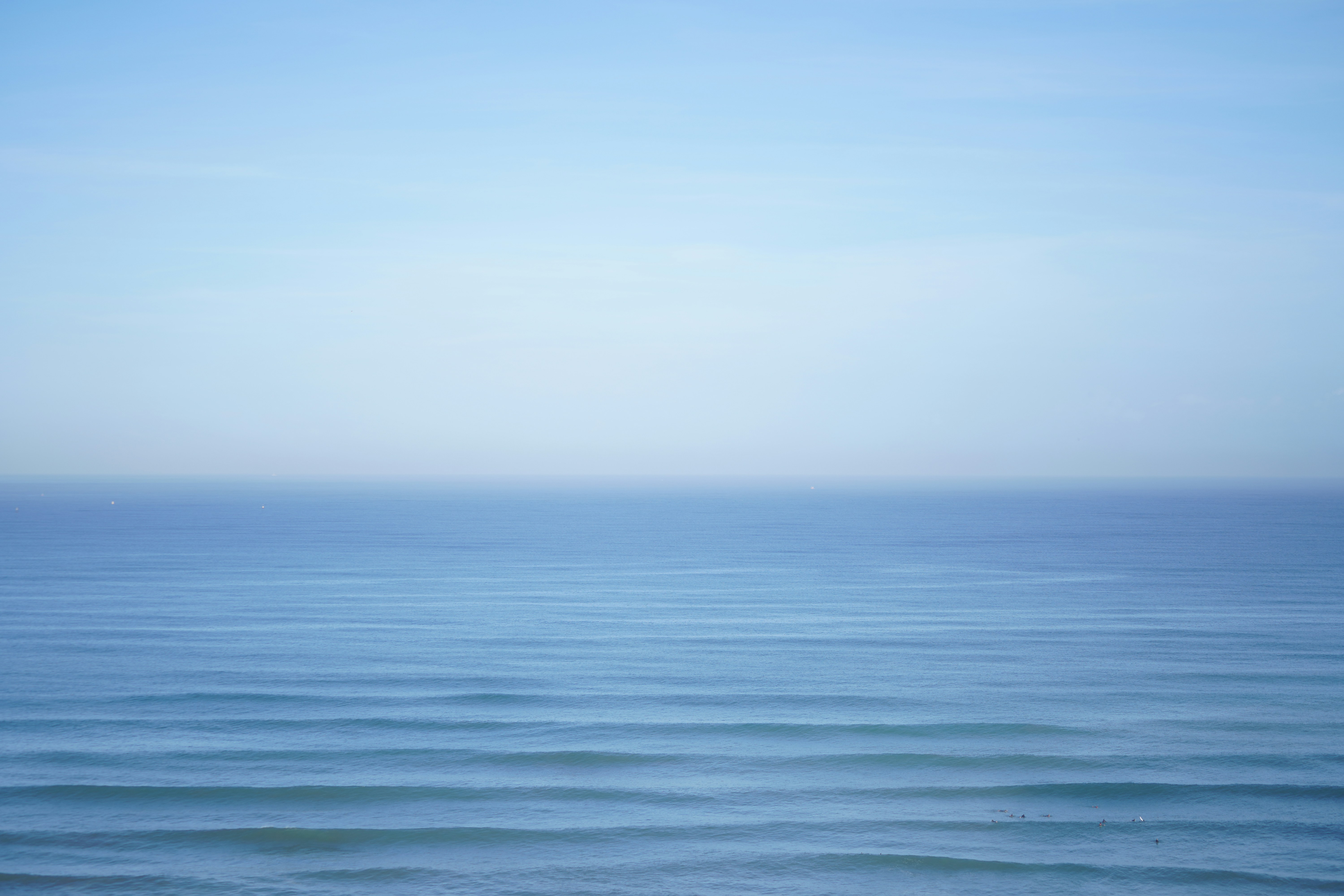 Vast blue ocean meeting a clear sky with gentle waves and distant surfers visible on the lower right.