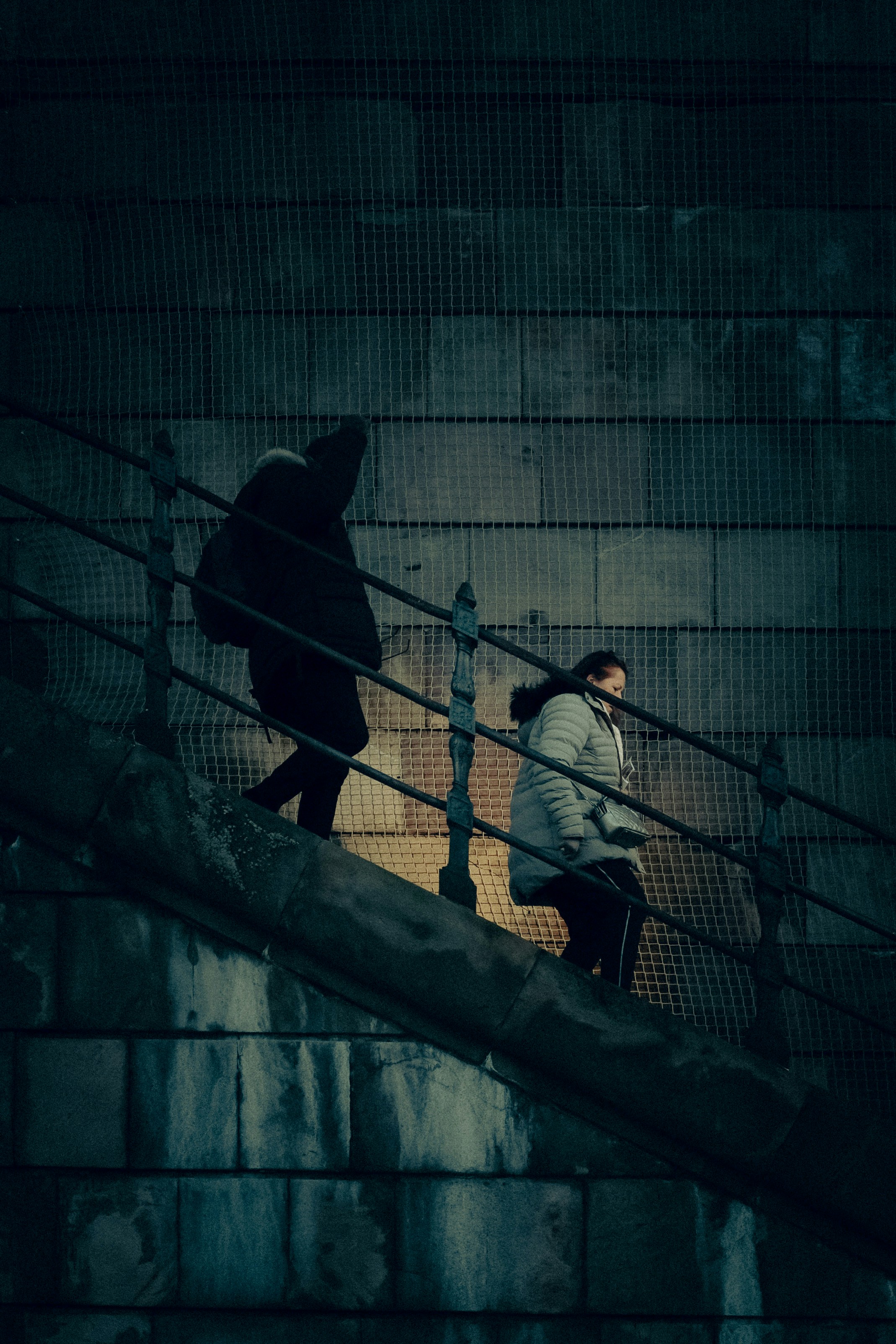 Teal-Lit Ascent: A pale-clad figure climbs a damp stairwell by a chain-link wall