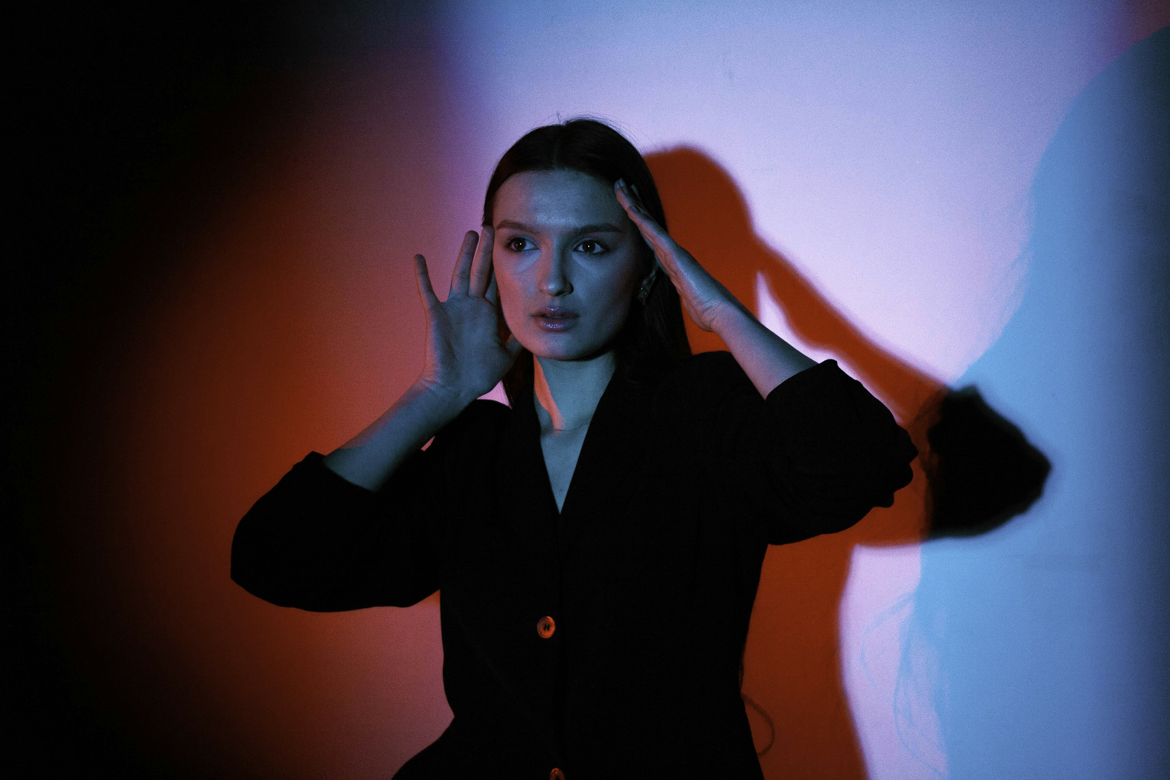 Person in a dark outfit poses with hands near face, illuminated by vibrant red and blue lighting.
