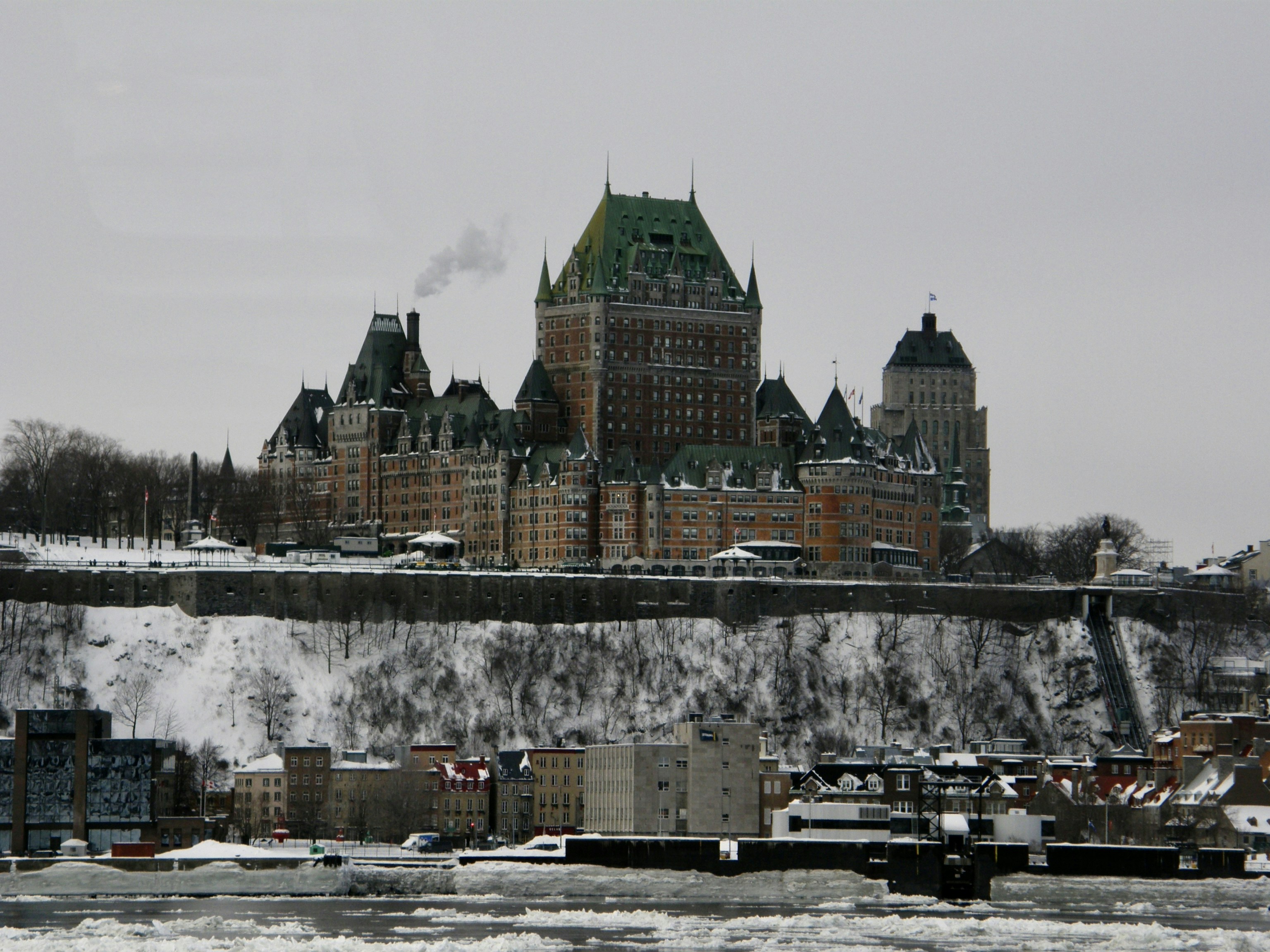 Historic castle perched on a snowy cliff, with smoke rising from a chimney against a gray sky.