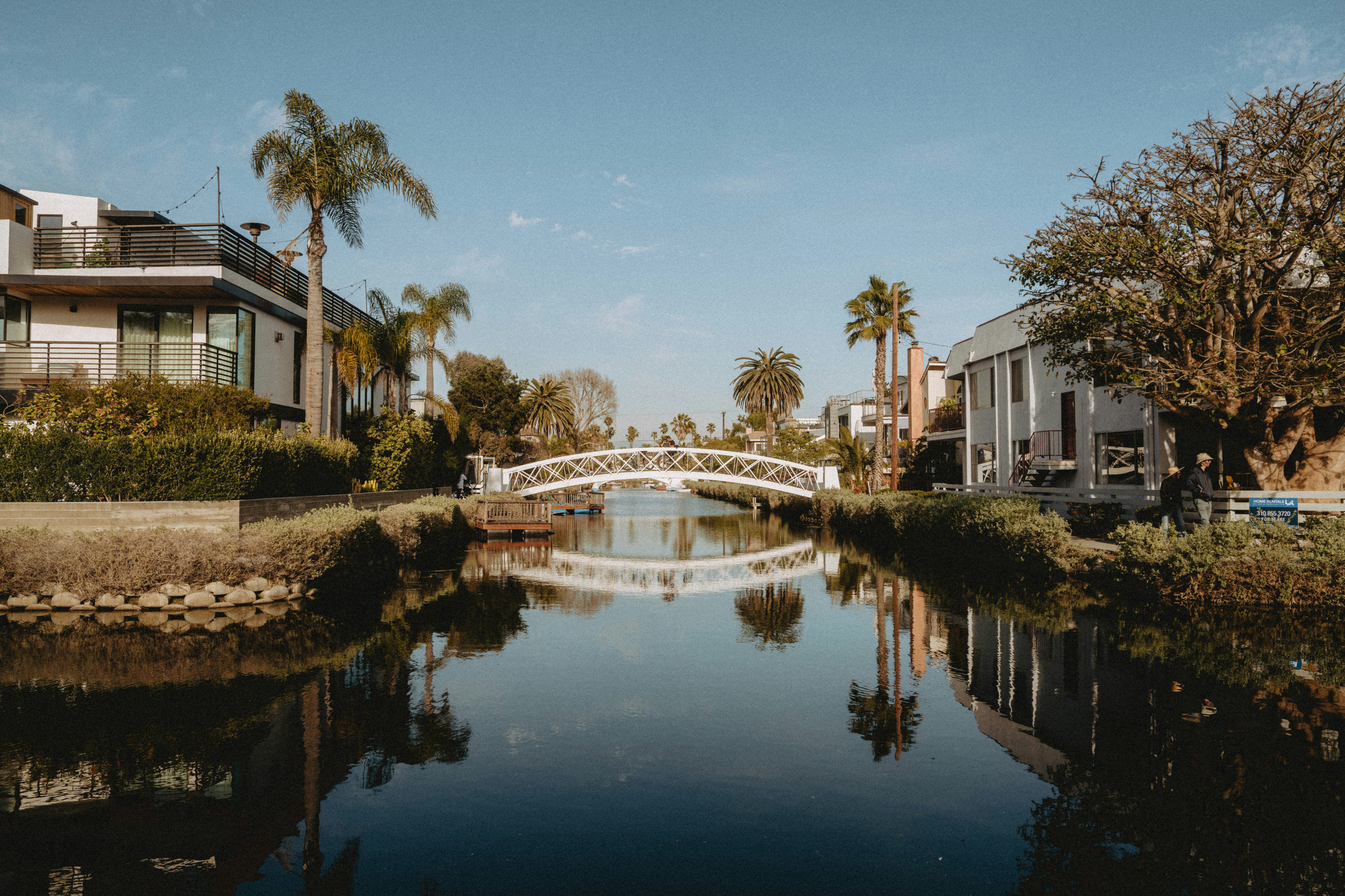 Calm canal waters reflecting a white bridge and surrounding palm trees under a clear blue sky.