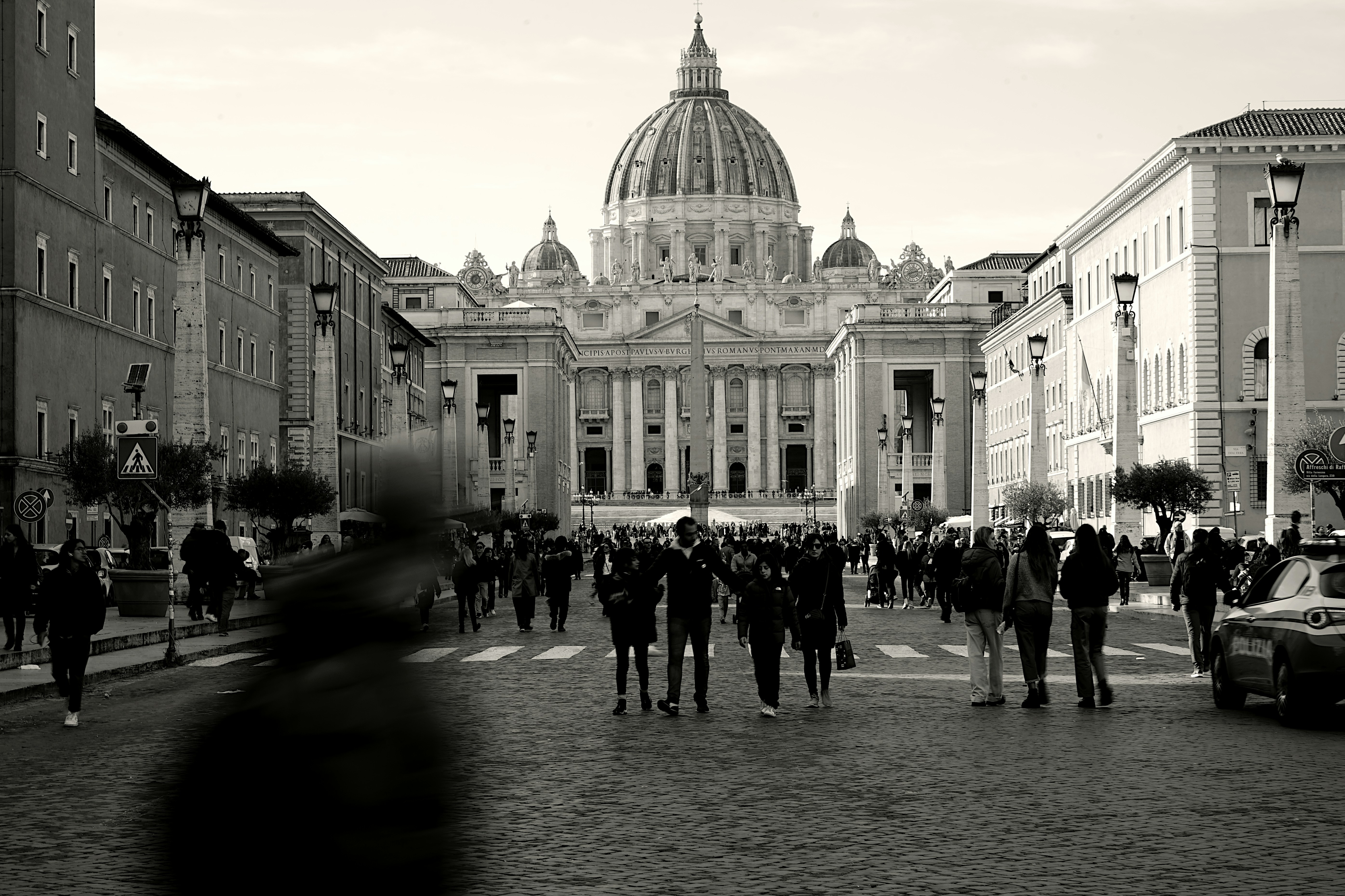 Crowd of visitors walking towards St. Peter's Basilica in Vatican City, framed by historic architecture. Black and white tones enhance the timeless atmosphere.